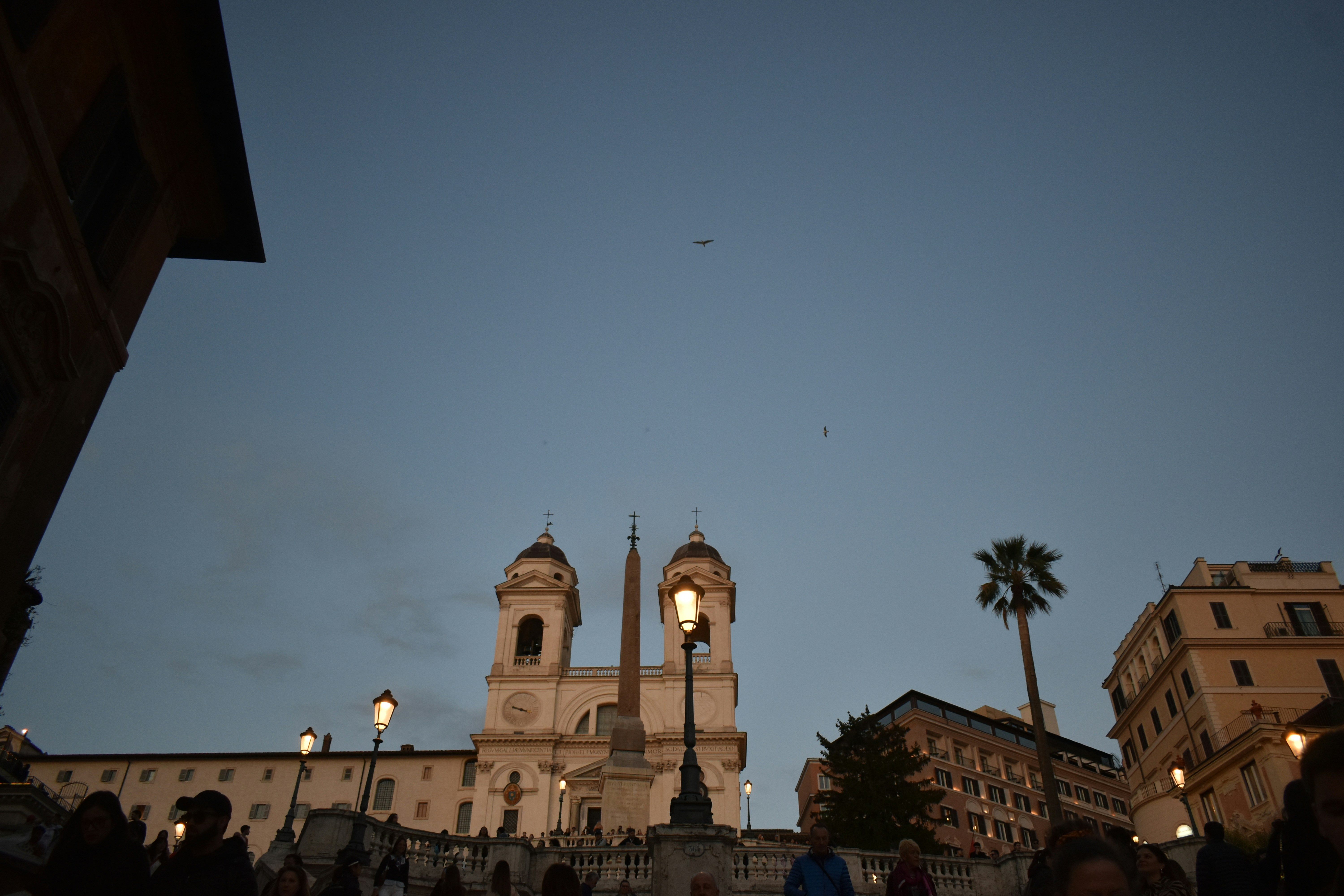 Twilight view of the Spanish Steps and Trinità dei Monti Church framed by surrounding buildings and a palm tree.