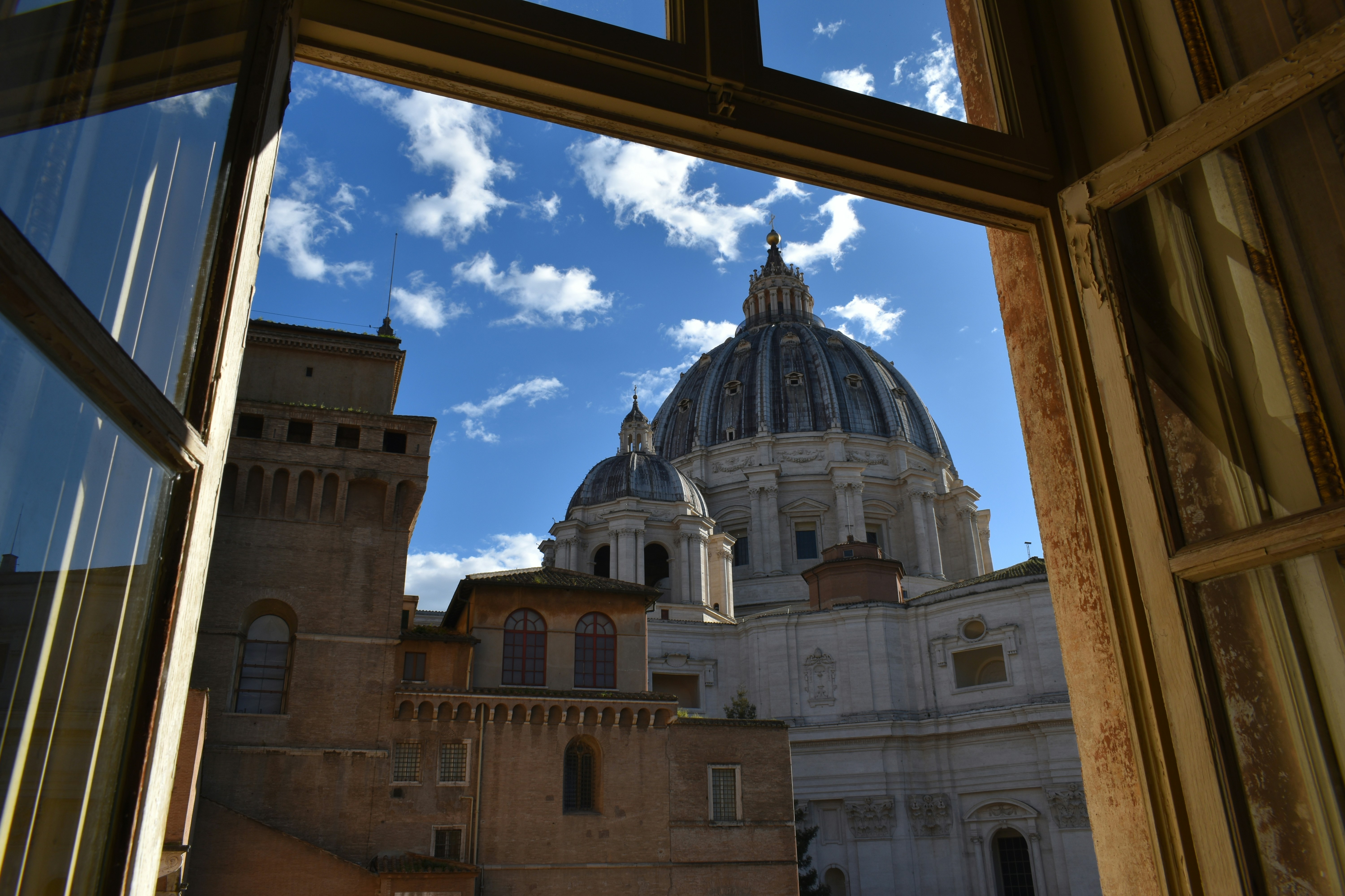 Saint Peter's Basilica dome viewed through an ornate window against a sky dotted with clouds.