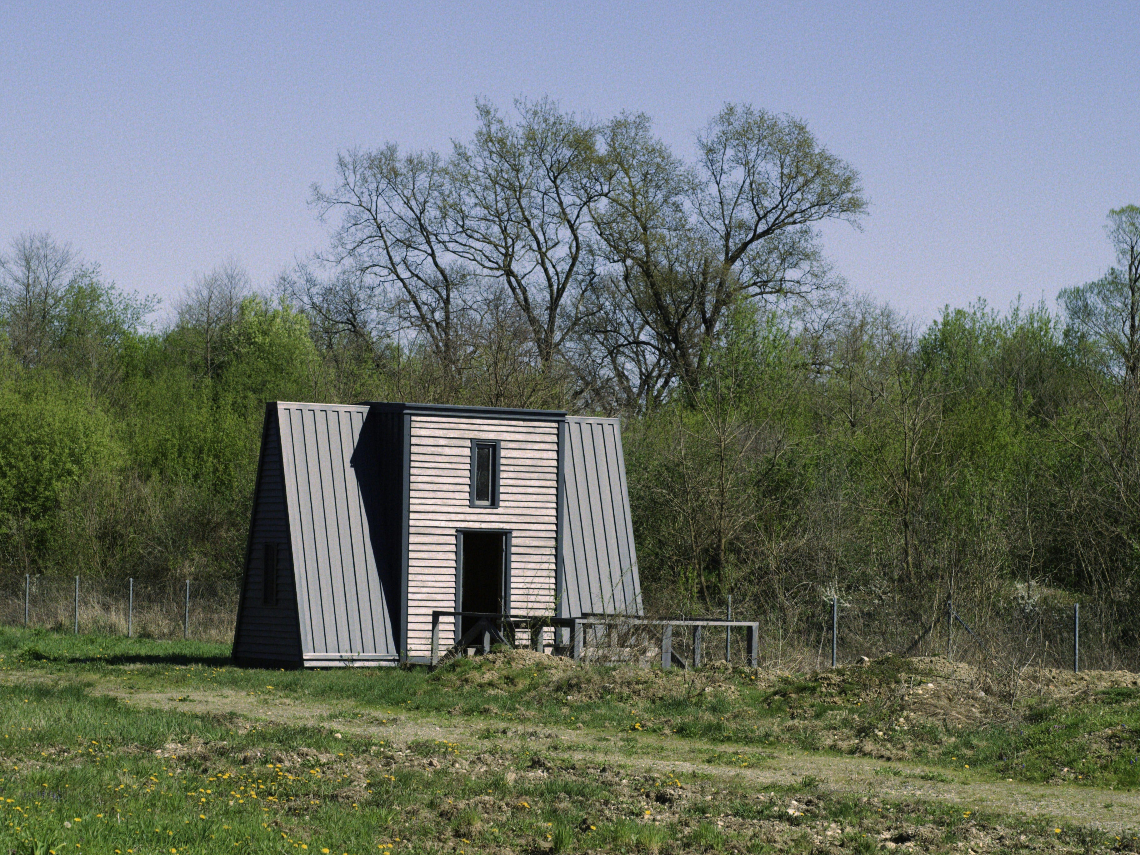Wooden A-frame cottage sits in a clearing surrounded by lush trees under a clear blue sky.
