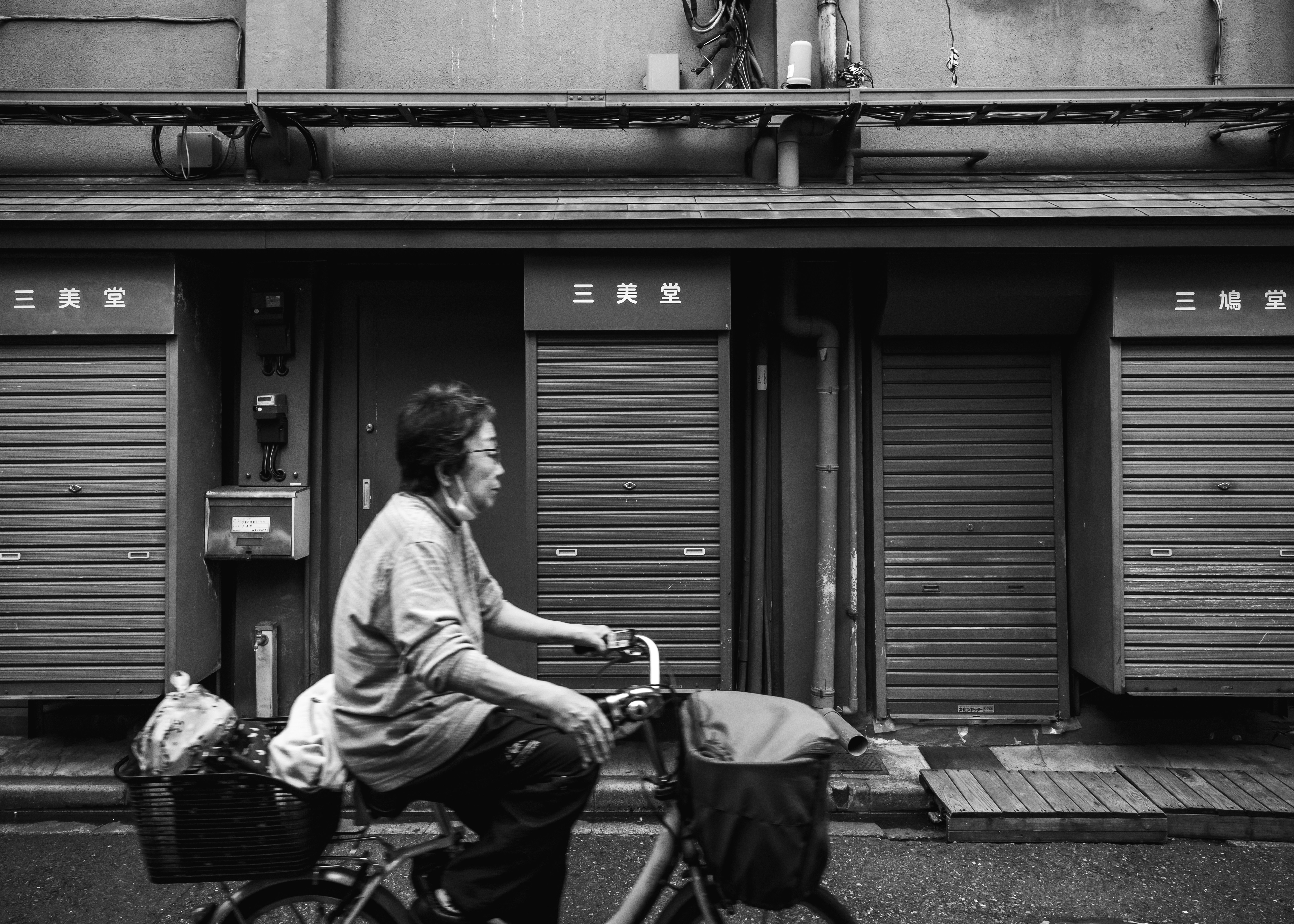 Black and white photo of a person cycling past closed storefronts on a quiet street.