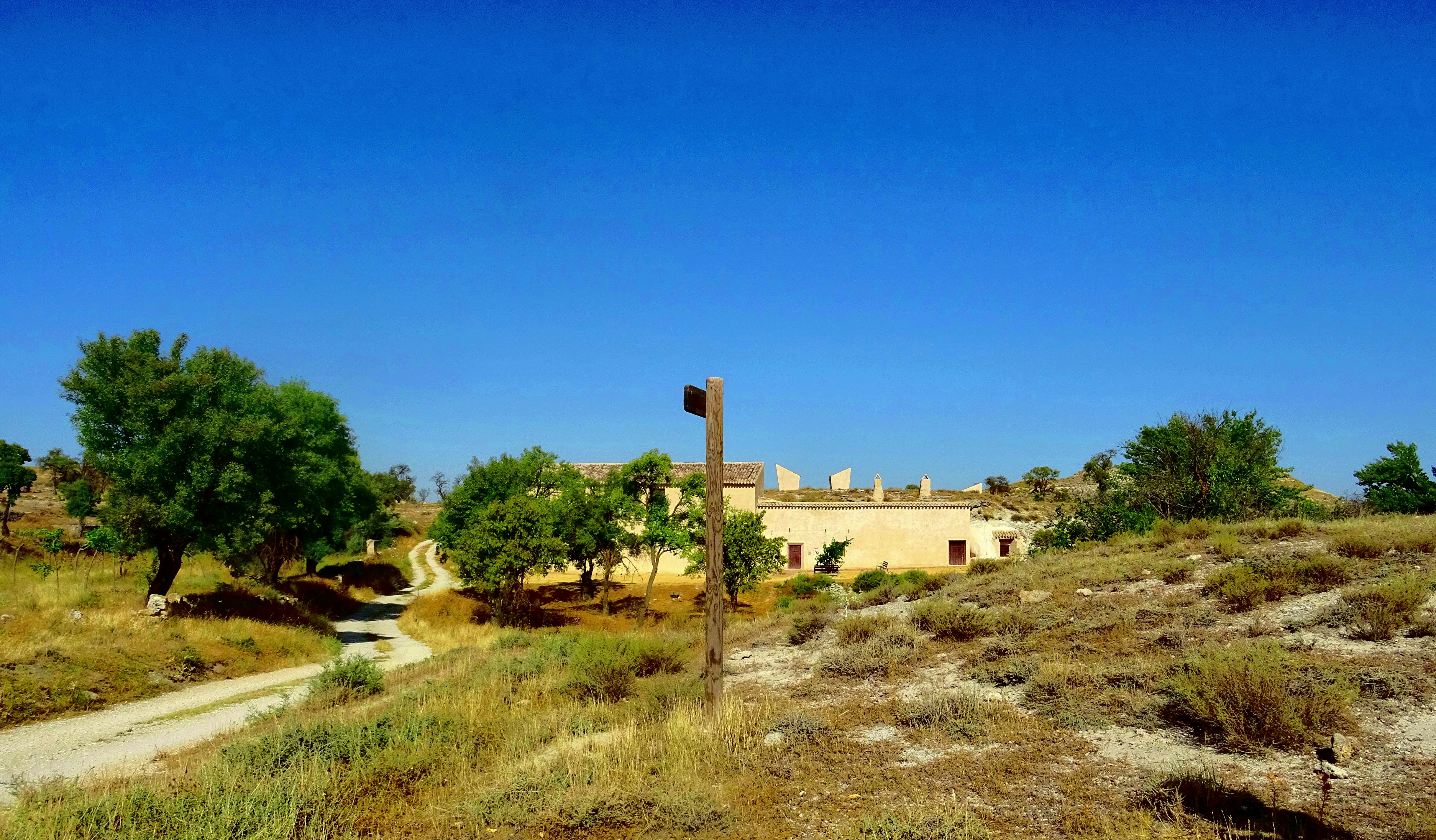 Historic building nestled in a sunlit, arid landscape with scattered greenery under a vibrant blue sky.