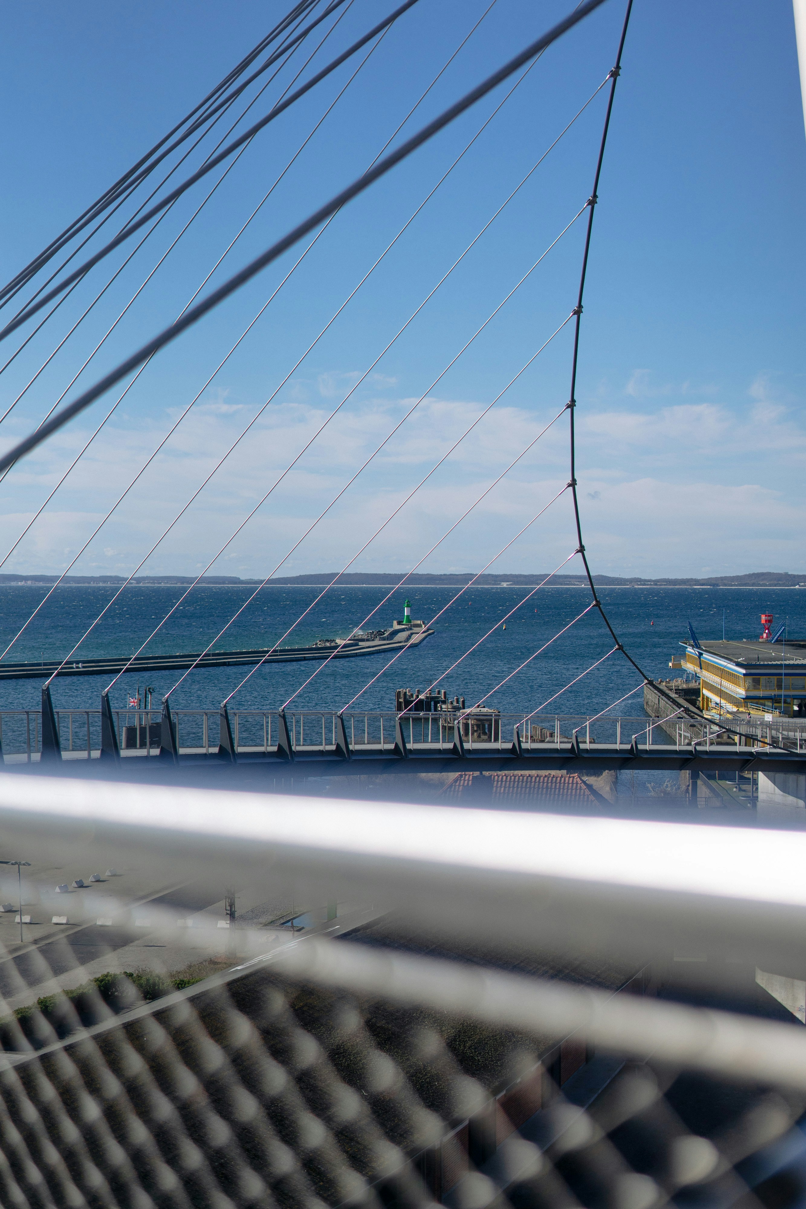 Steel cables of a suspension bridge frame a distant view of the ocean under a clear blue sky.