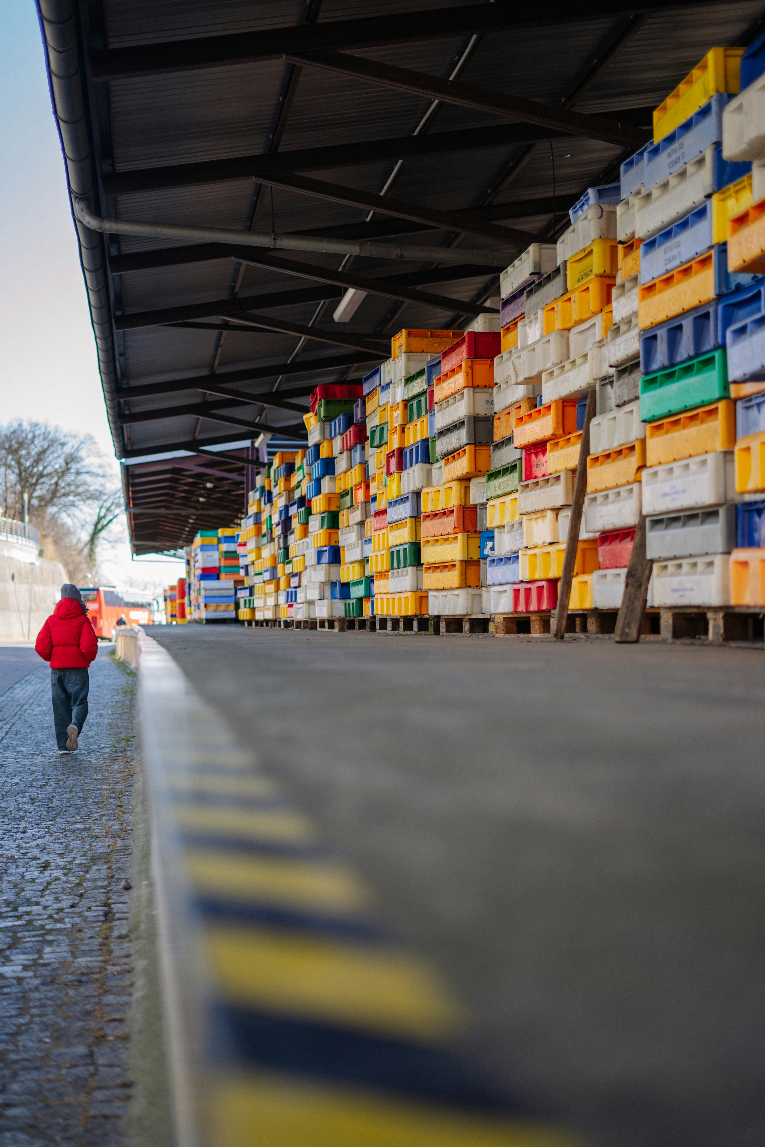 Stacks of brightly colored crates lined under a warehouse roof along a cobblestone path as a person walks beside them.