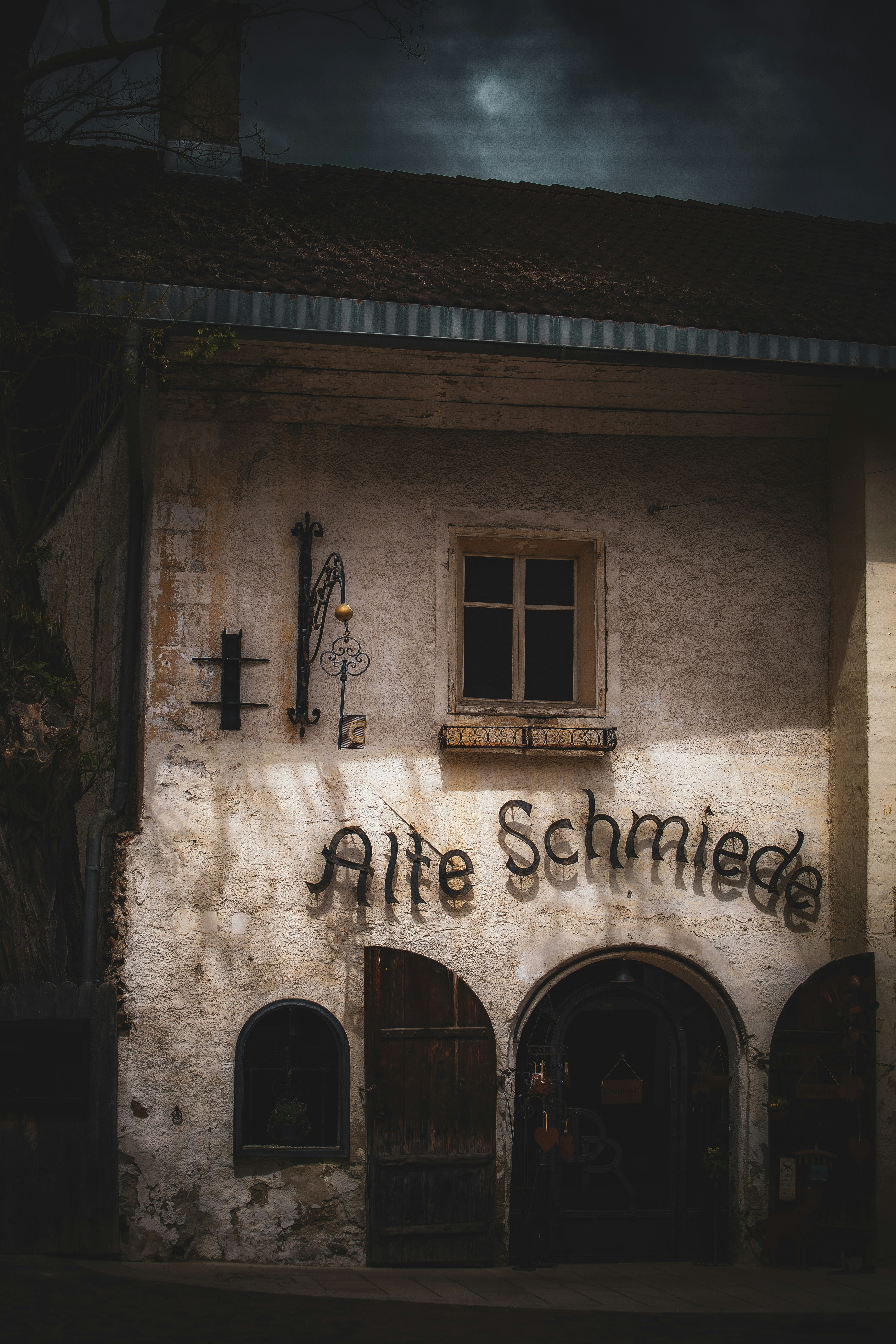 Rustic building with 'Alte Schmiede' sign, shadowed under dramatic dark clouds.