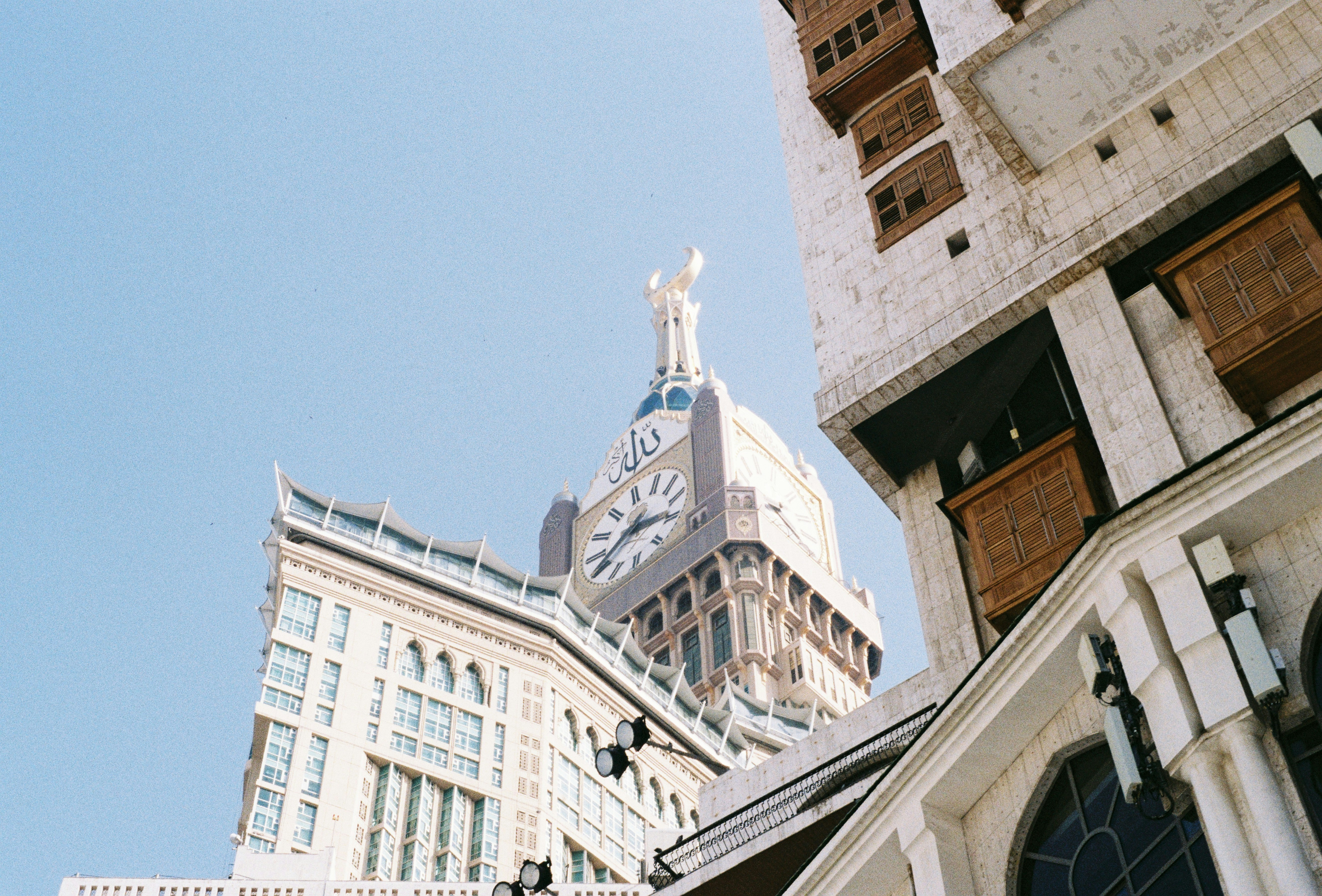 An angled cityscape featuring a prominent clock tower rising between modern and classical façades against a pale blue sky.