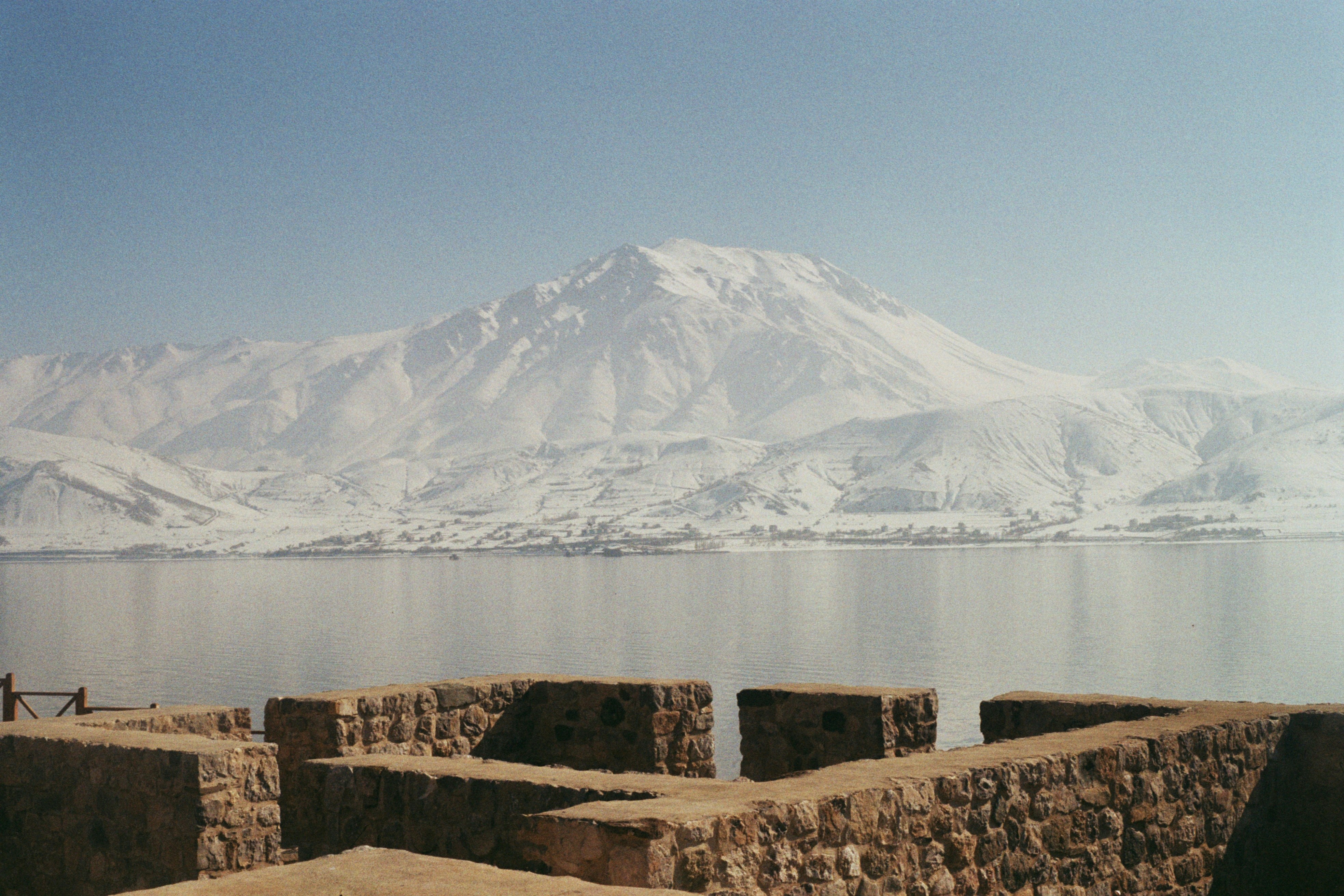 Snow-covered mountains rise majestically above a tranquil lake, framed by rugged stone structures in the foreground.