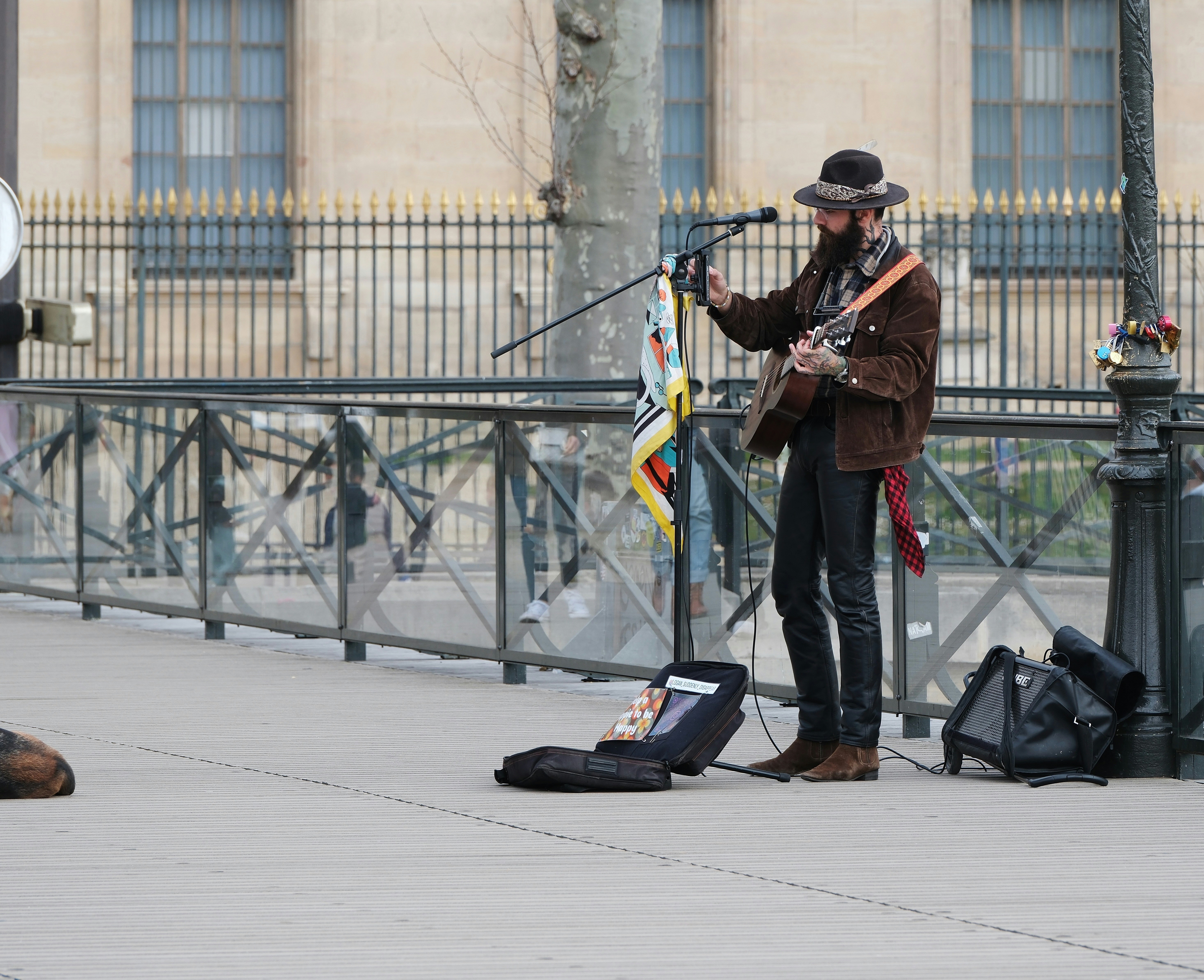 Street musician performing with a guitar and microphone near a fenced urban backdrop.