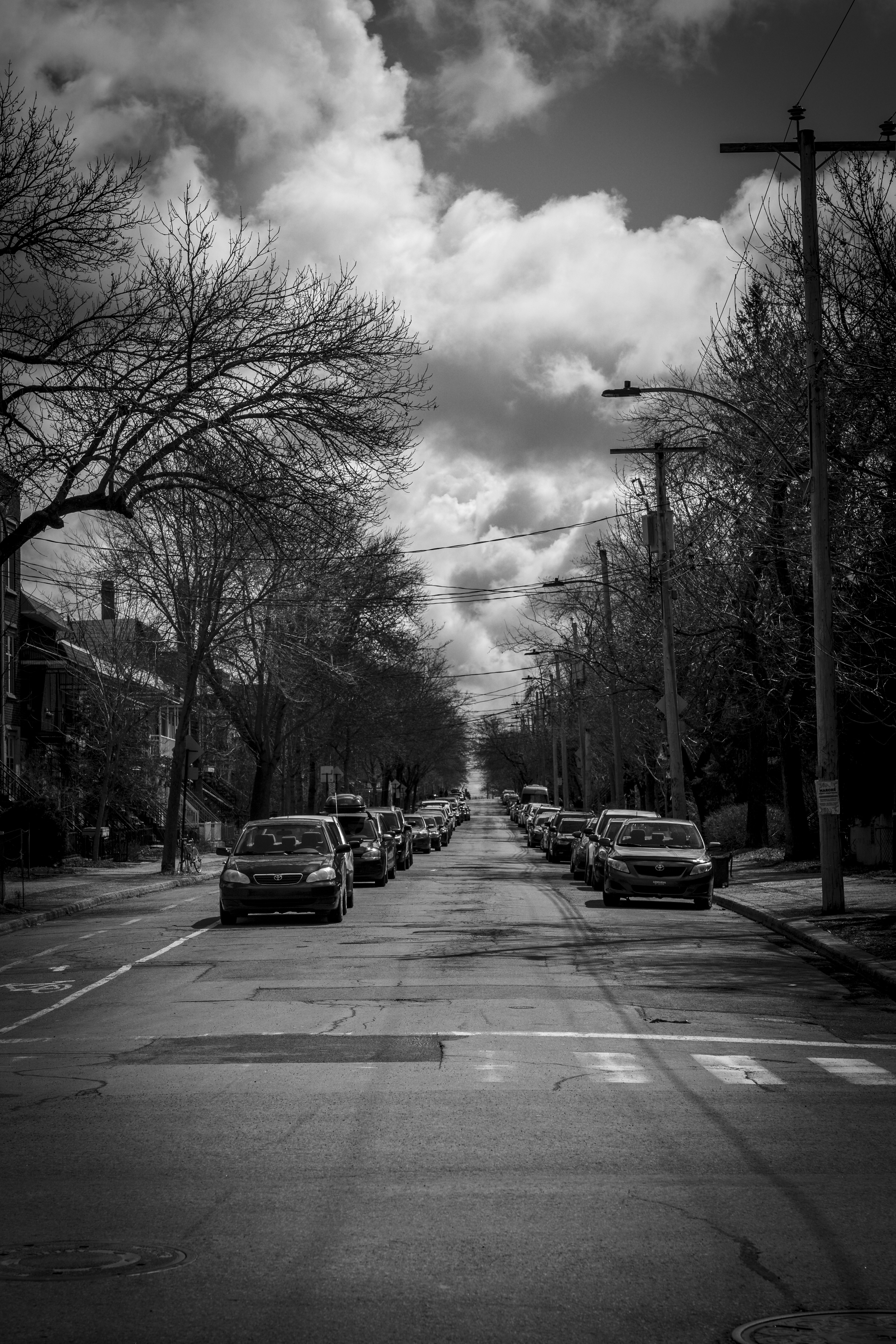 Black and white street scene with parked cars and overcast sky, creating a dramatic perspective.