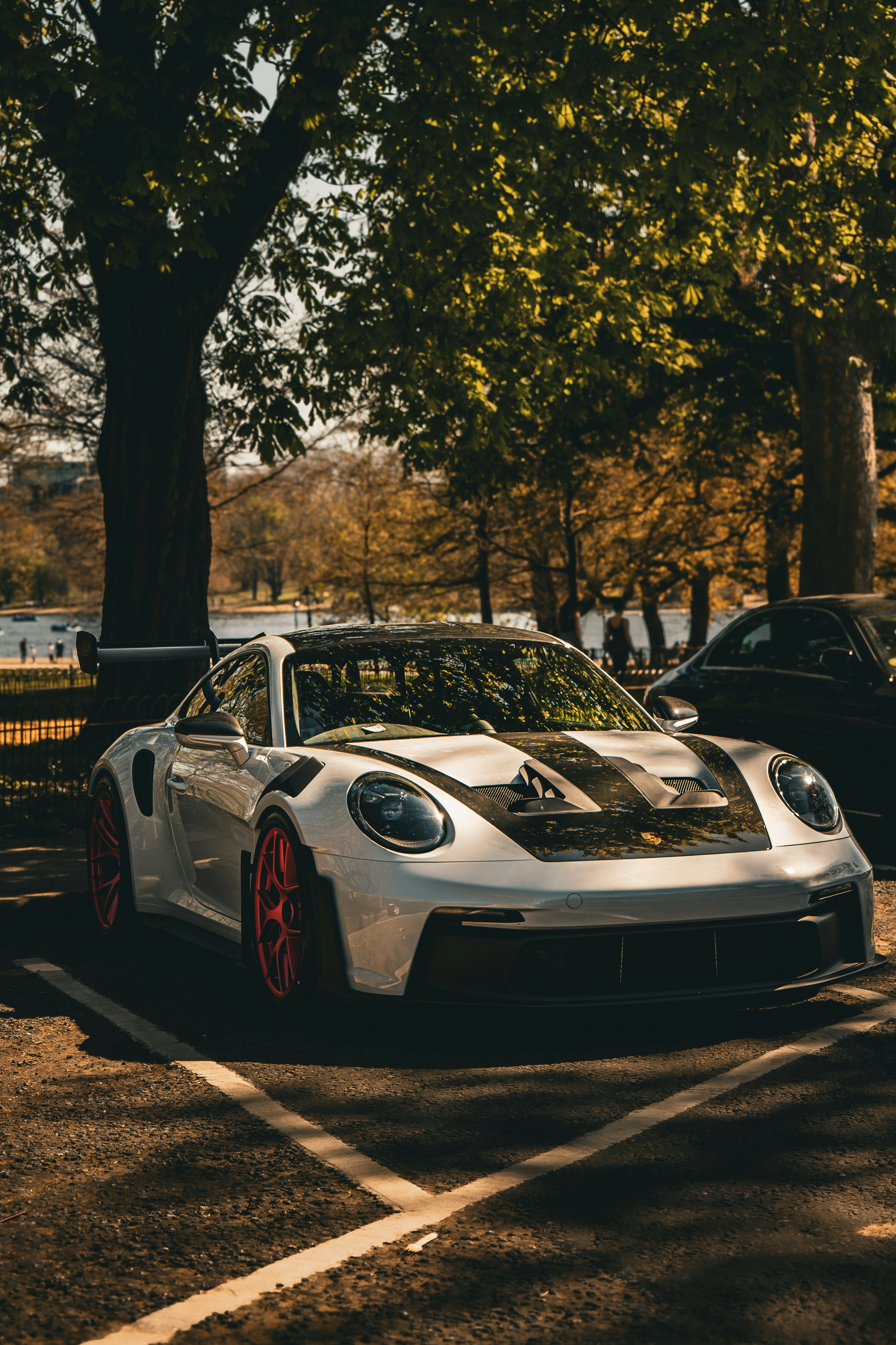Sleek white Porsche 911 GT3 RS with red wheels, parked under tree canopy in sunlit setting.