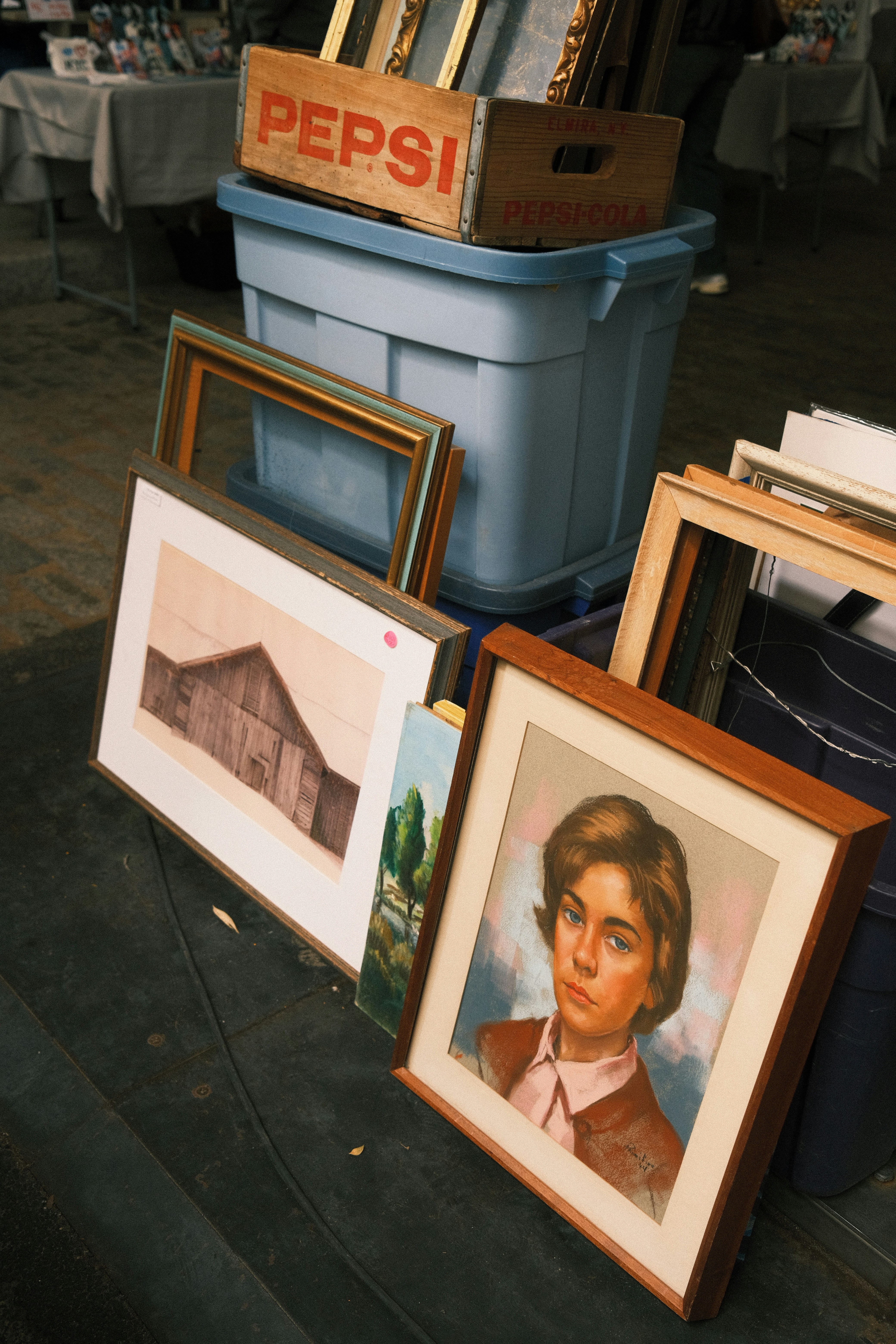 A collection of framed artworks and photographs displayed at a market, featuring a portrait of a young girl in a vintage style. The scene captures the nostalgia of artistic treasures.