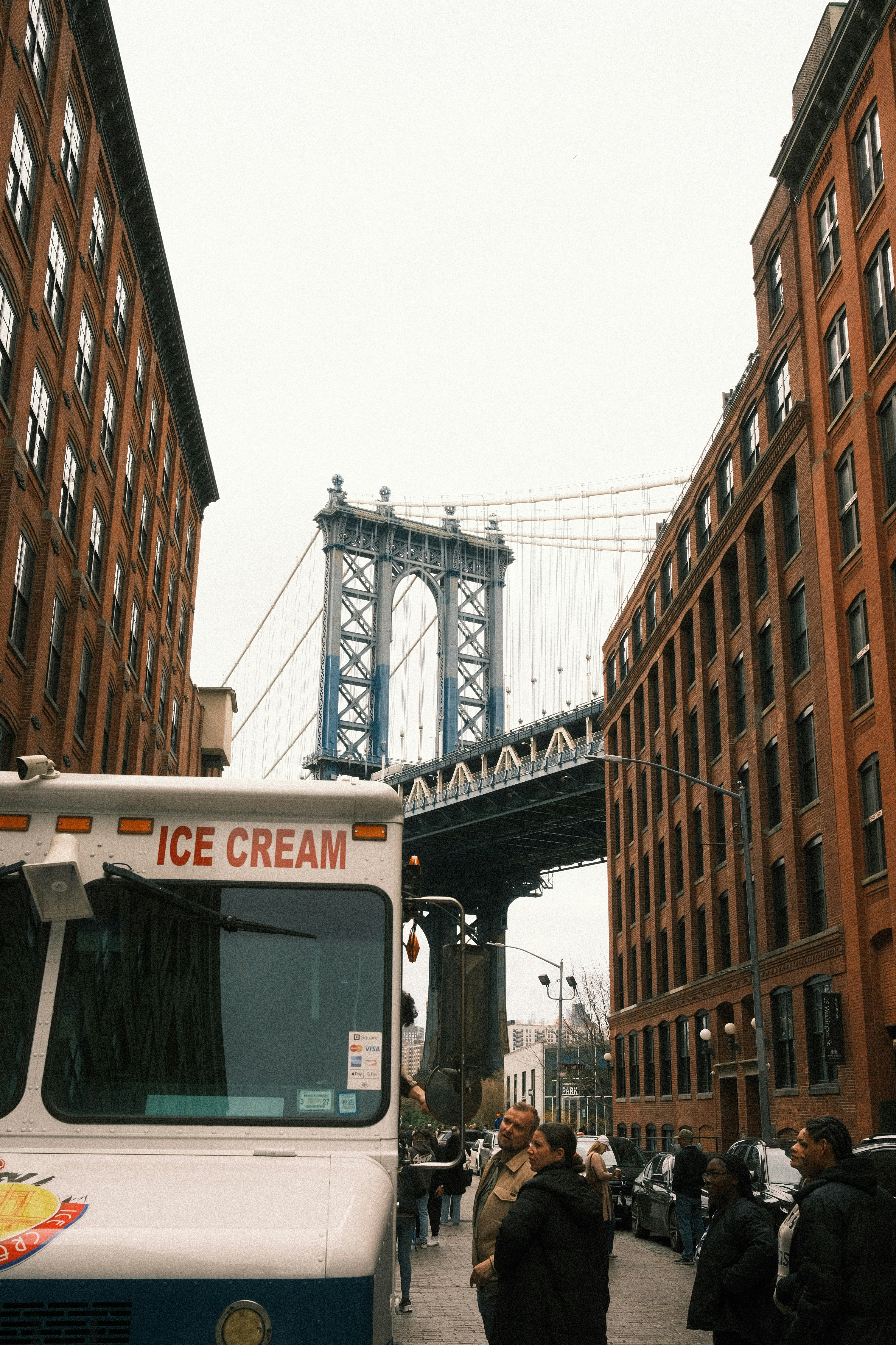 Street scene with ice cream truck framed by brick buildings and a bridge in the background.
