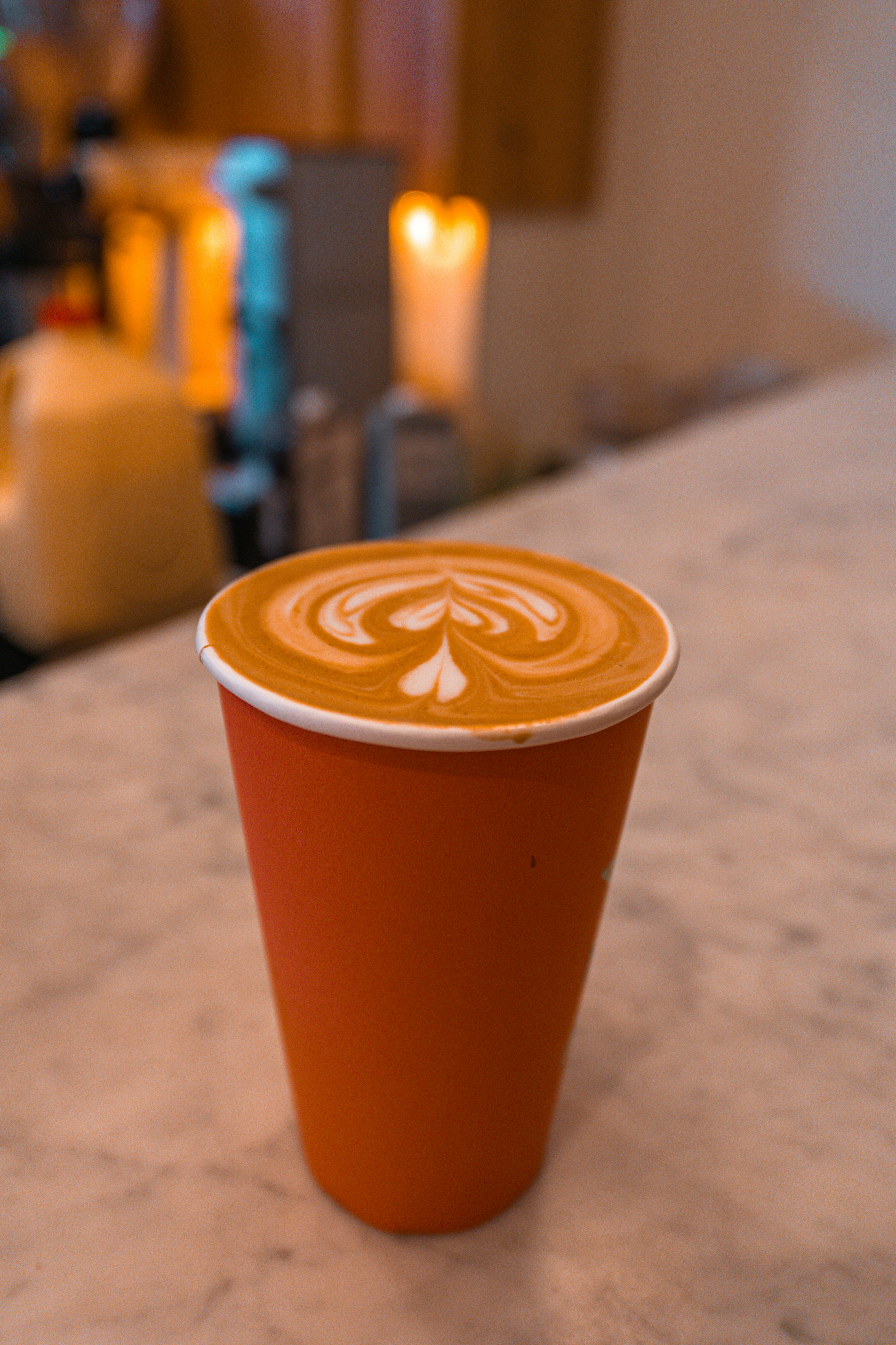 Orange cup of latte with heart-shaped foam design on a marble countertop.