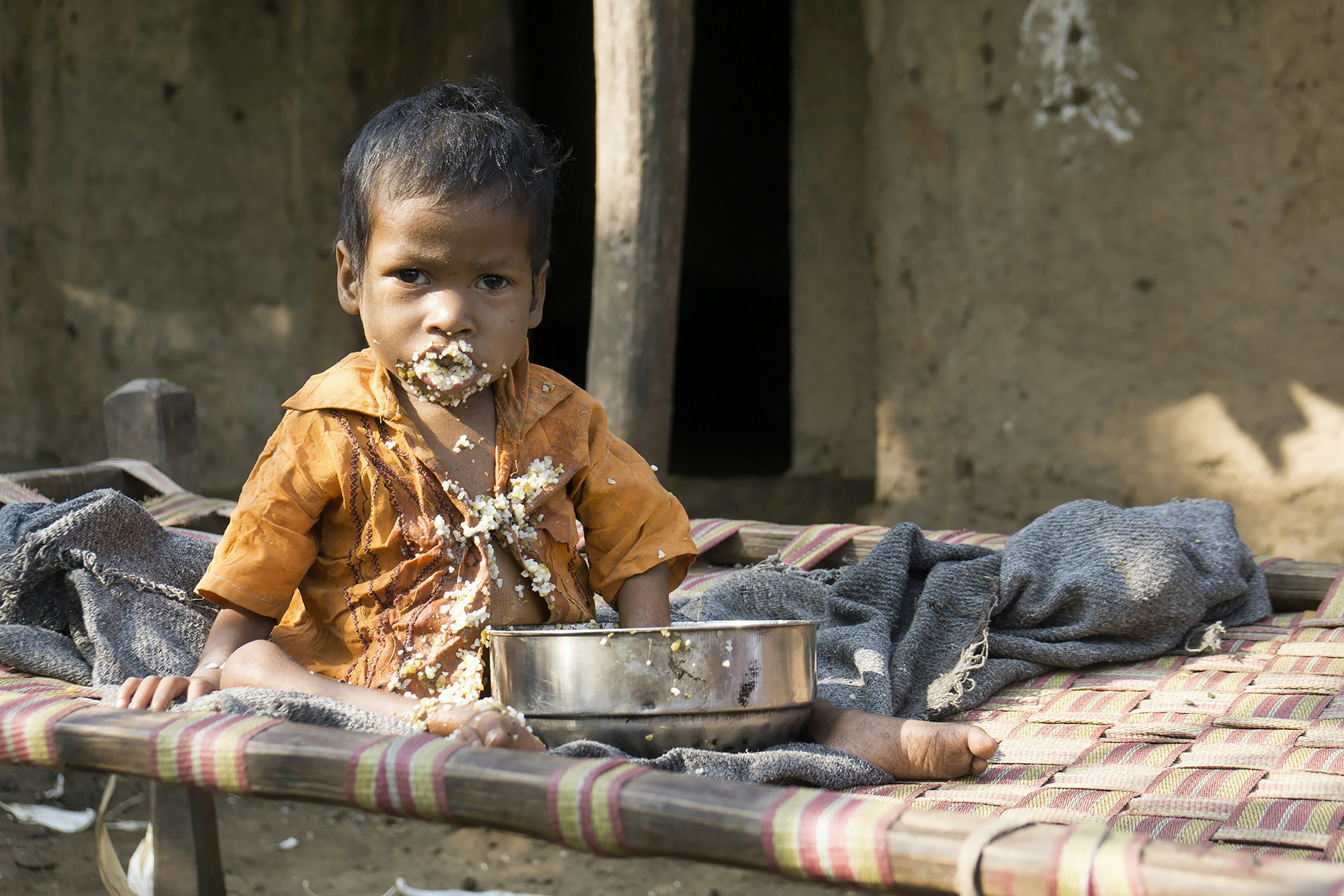 Young child sitting on a woven cot, eating from a metal bowl with a dirt wall backdrop.