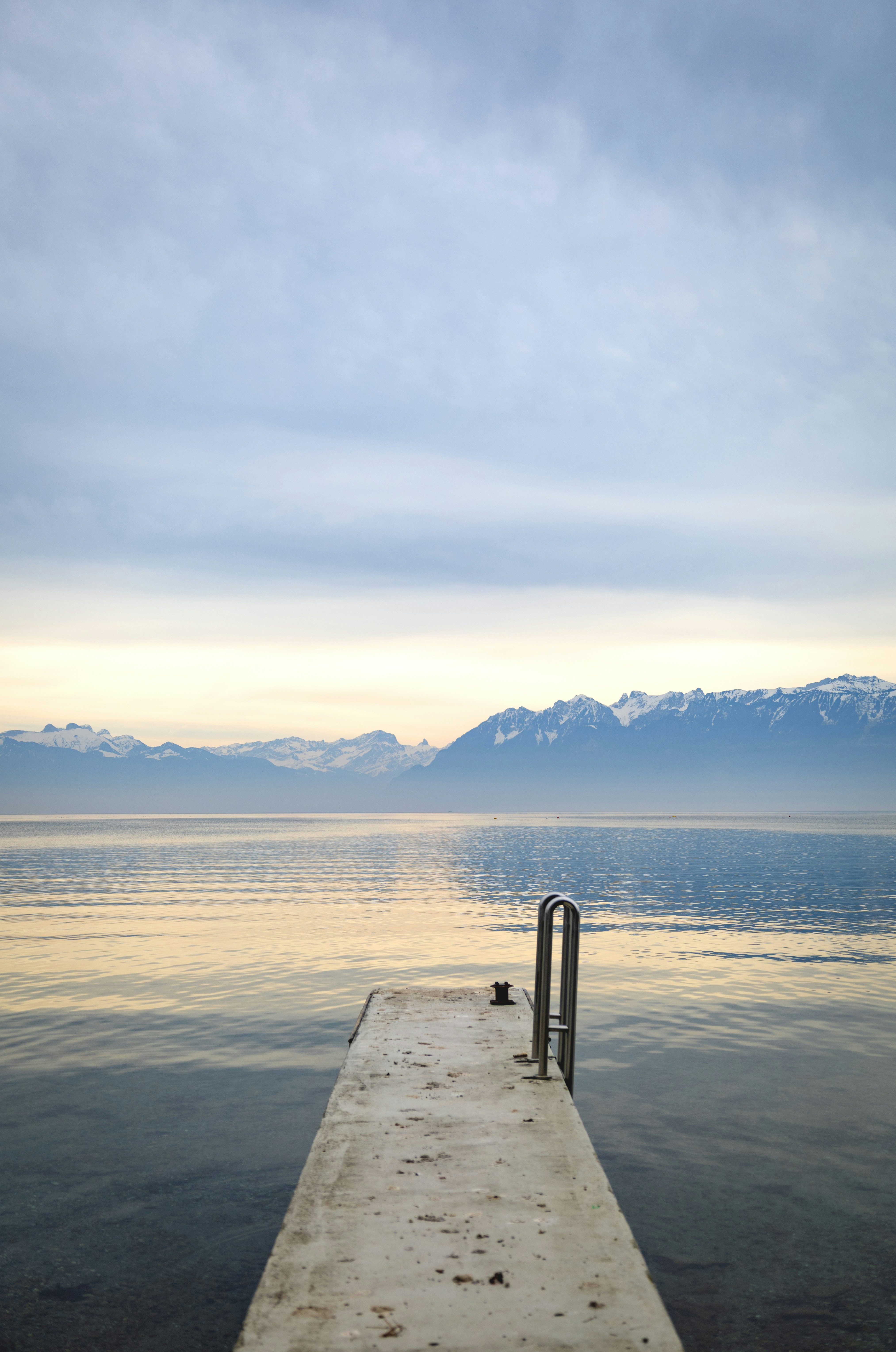 Concrete pier extending into serene lake with distant mountains under a pastel sky.