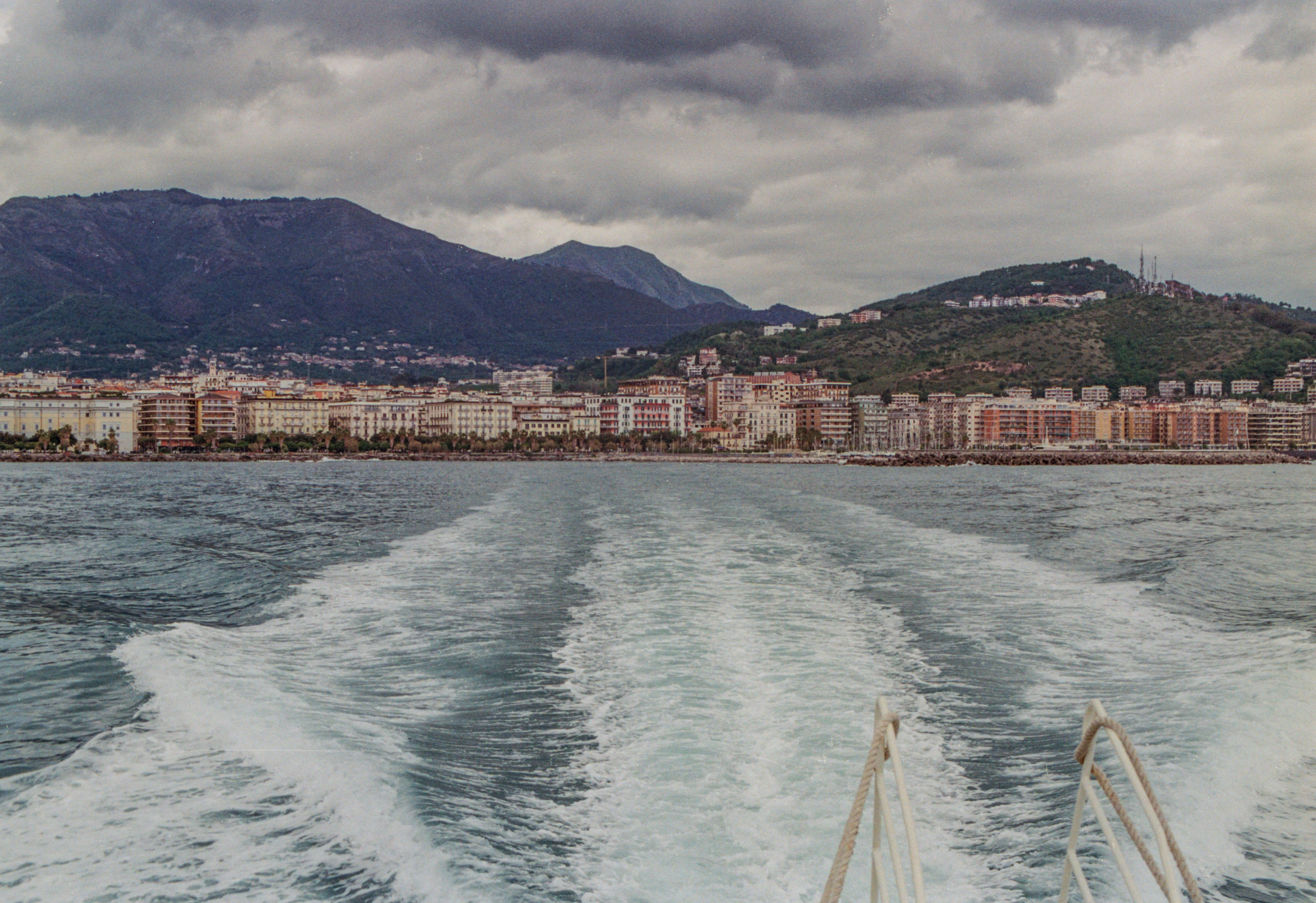 Foamy boat wake leads to a distant coastal town under a cloudy sky, with mountains in the background.