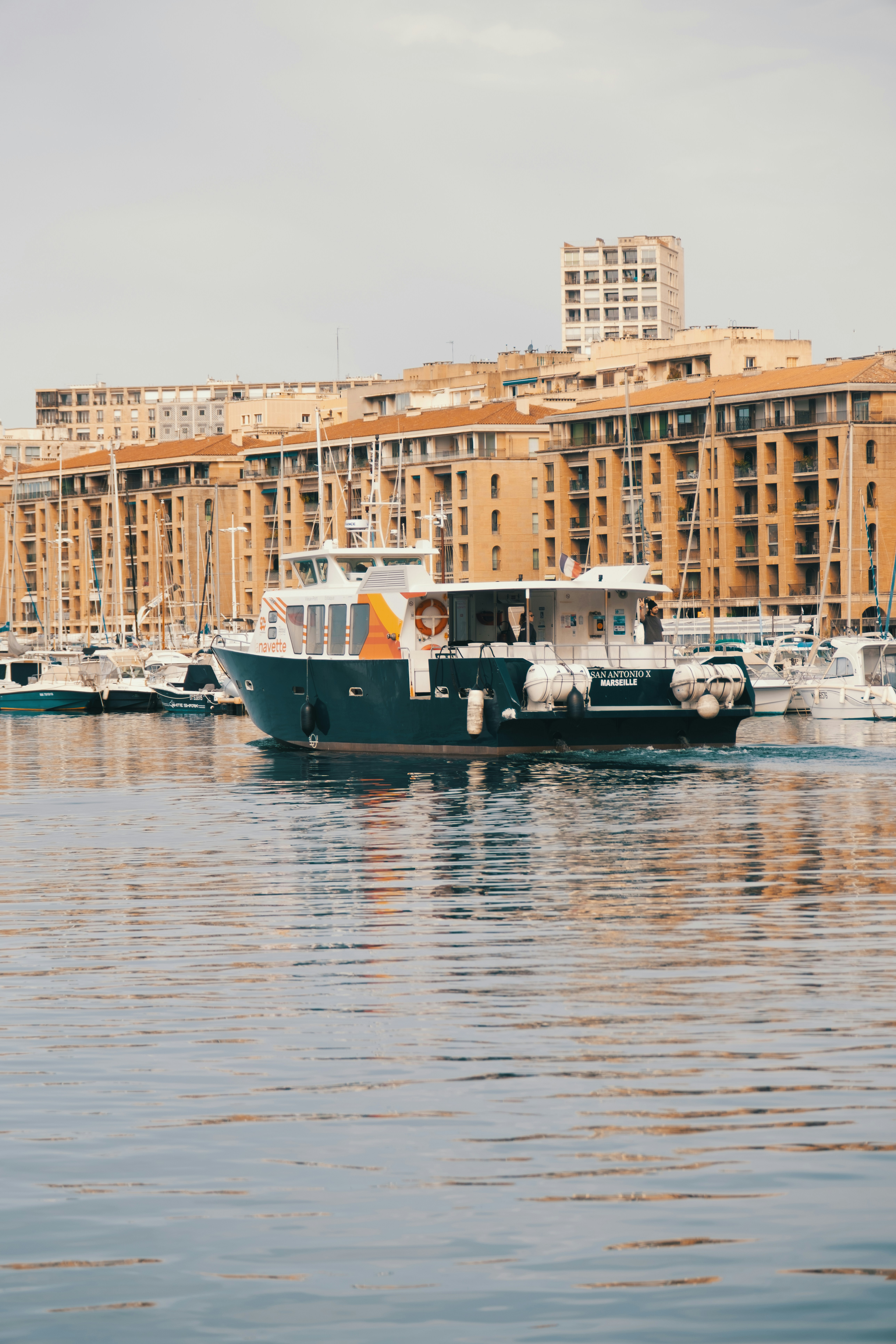 Boat gliding on calm waters in front of a row of yellow-toned buildings under a cloudy sky.
