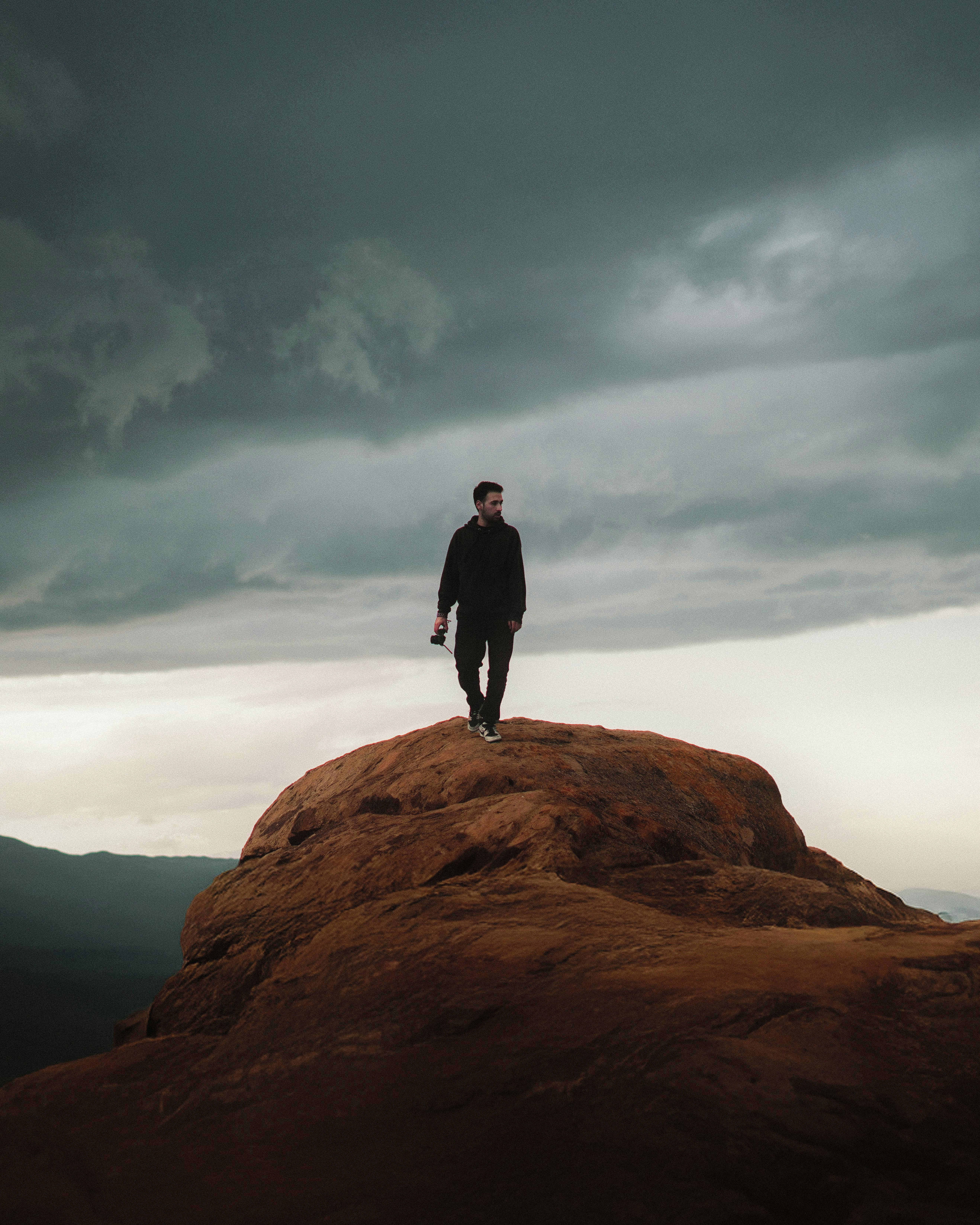 Person standing atop a rocky hill under dramatic, cloud-filled skies.
