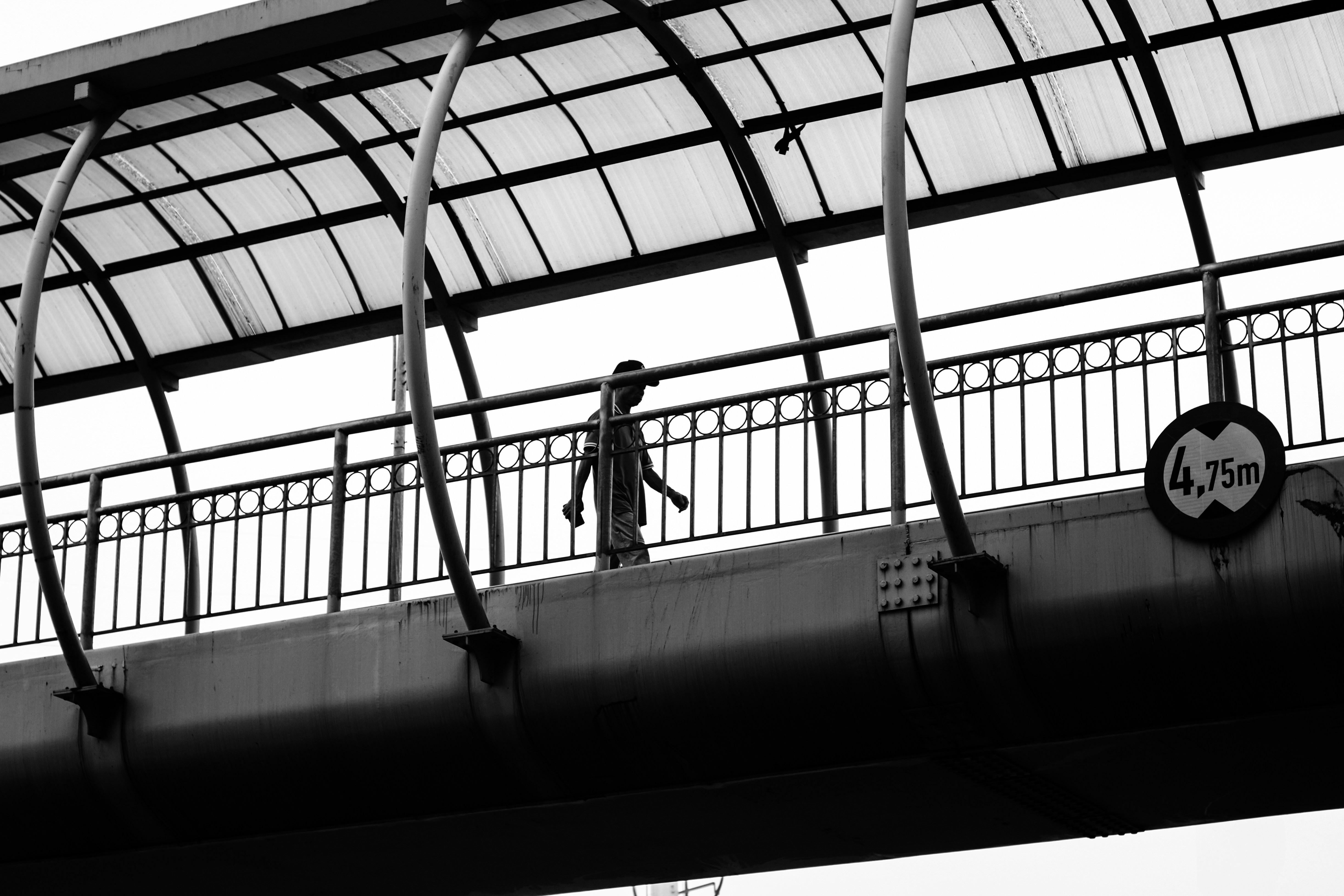 Person walking across a covered pedestrian bridge with curved metal and glass structure in monochrome.