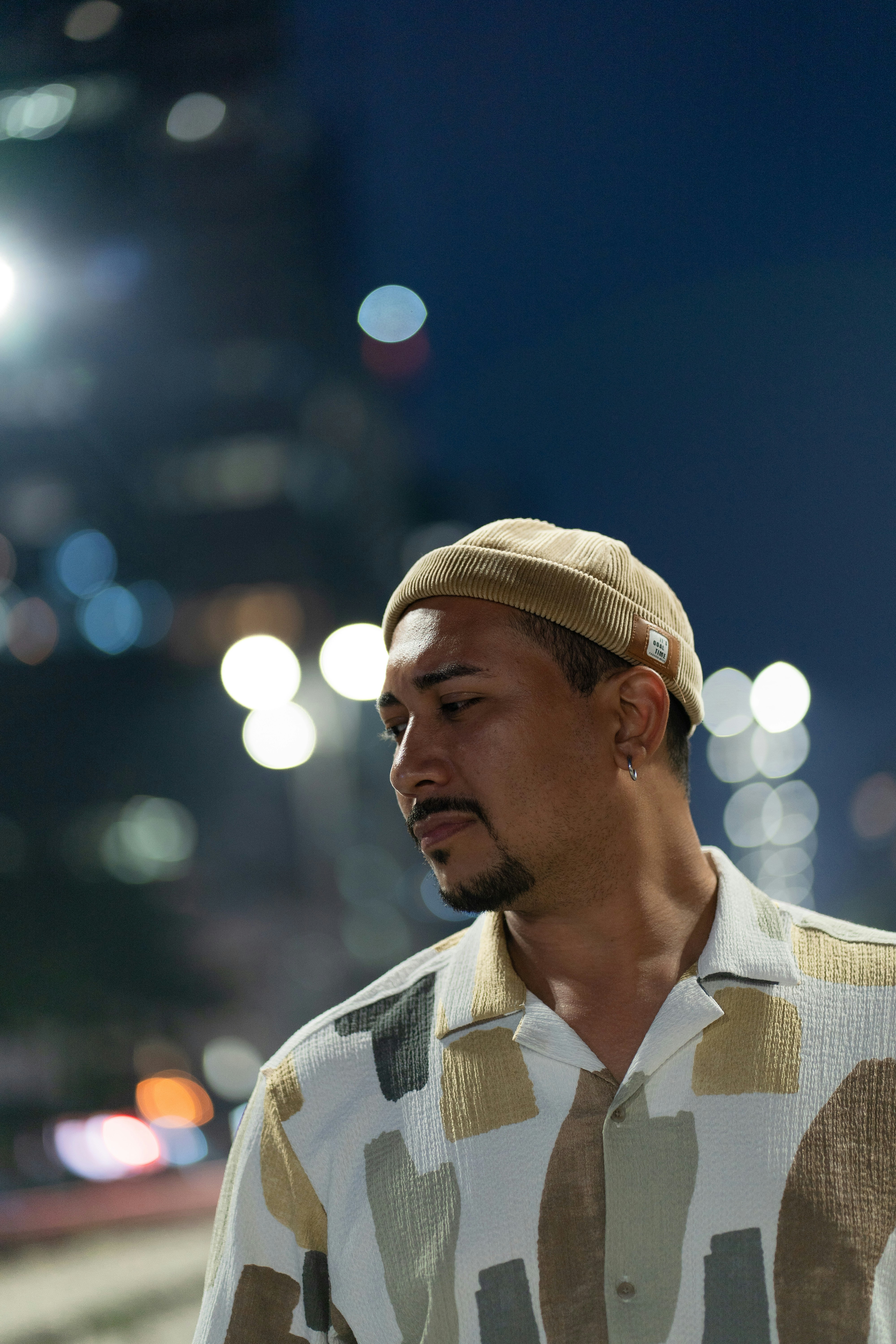 Man in patterned shirt gazing downward on a city street at night, surrounded by bokeh lights.