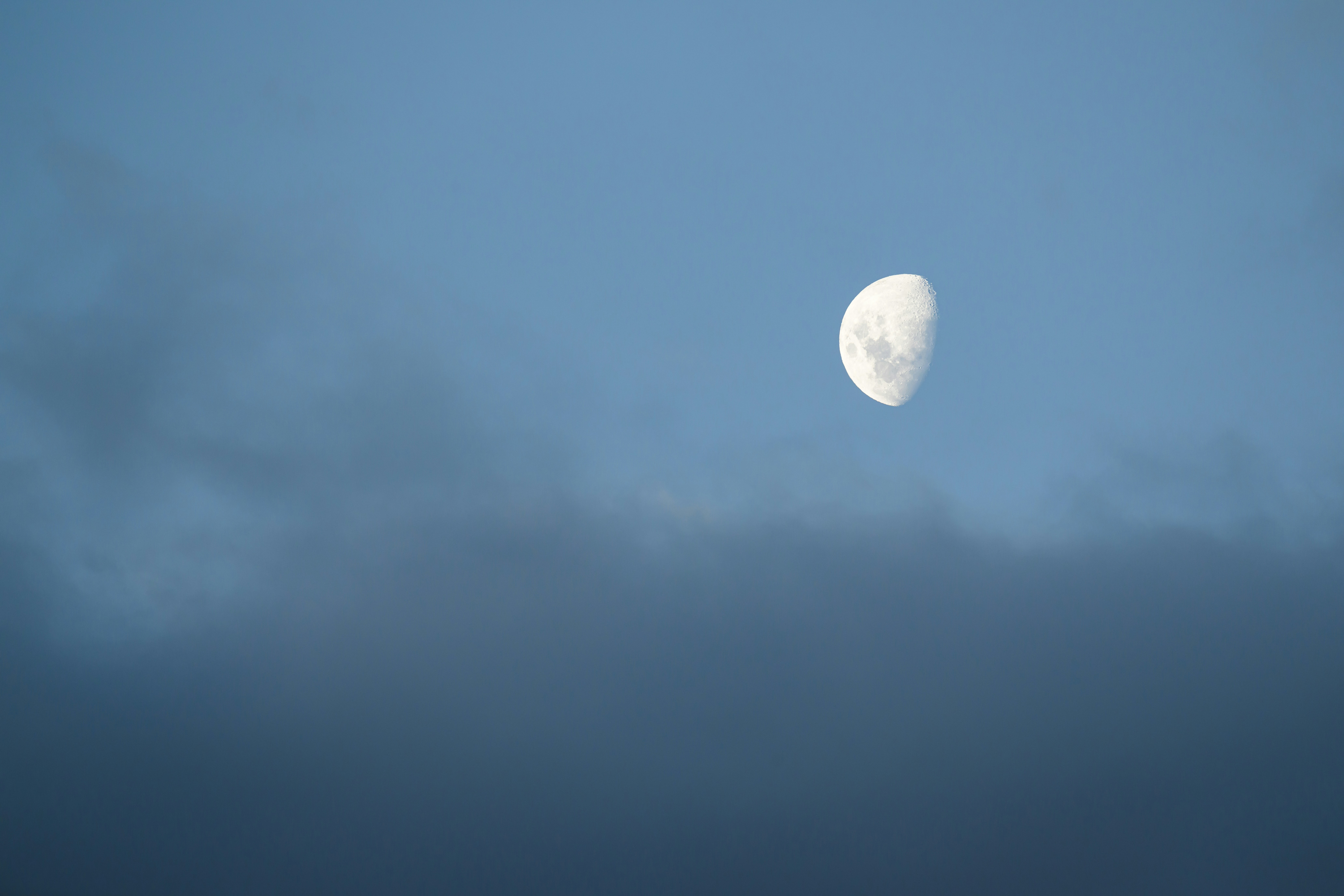 Half moon glows above a layer of dark clouds under a twilight sky.