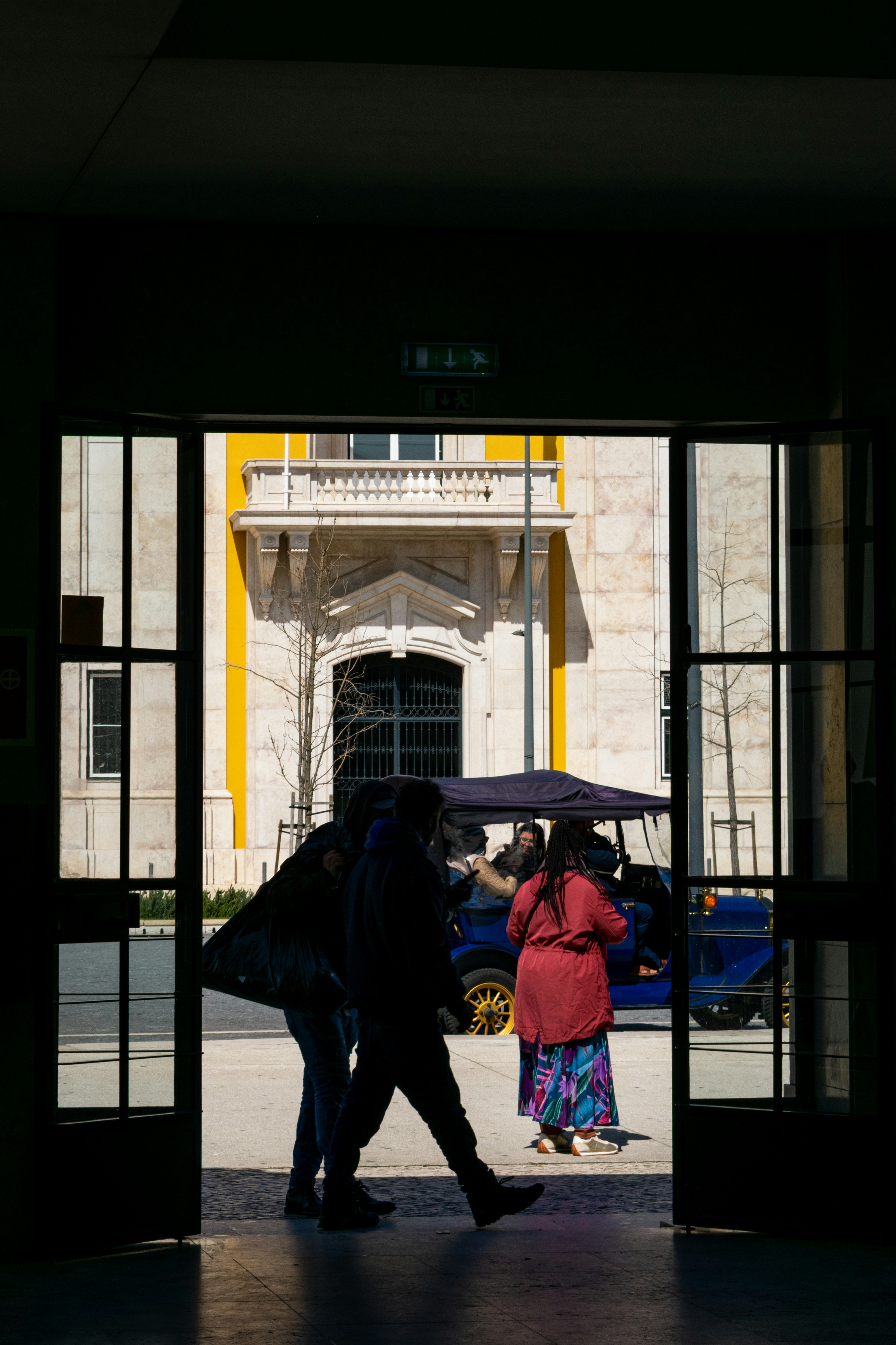 Silhouetted figures walk through an open doorway, revealing a sunlit street scene outside.