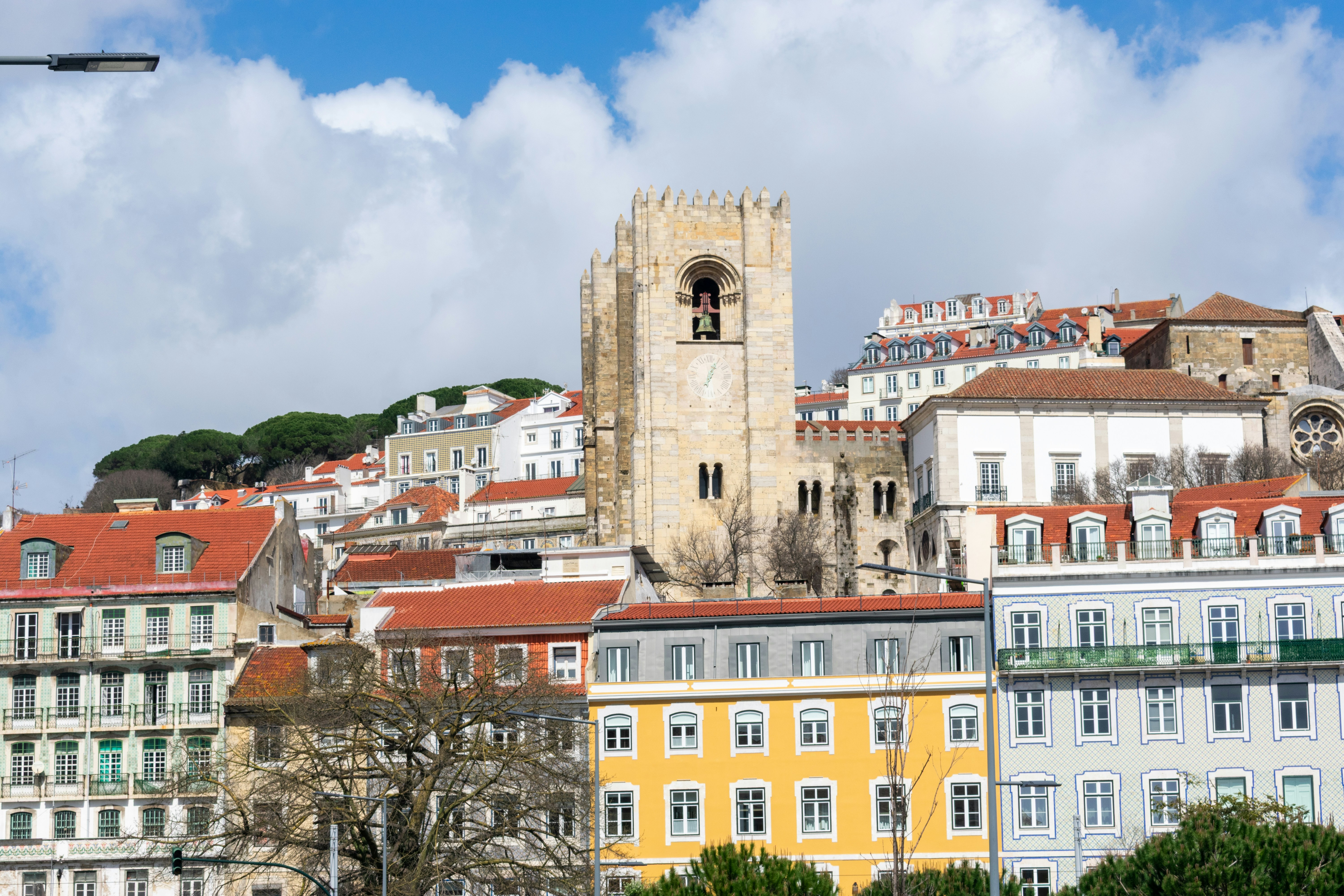 Lisbon's colorful hillside architecture crowned by an ancient stone tower under a bright blue sky with scattered clouds.