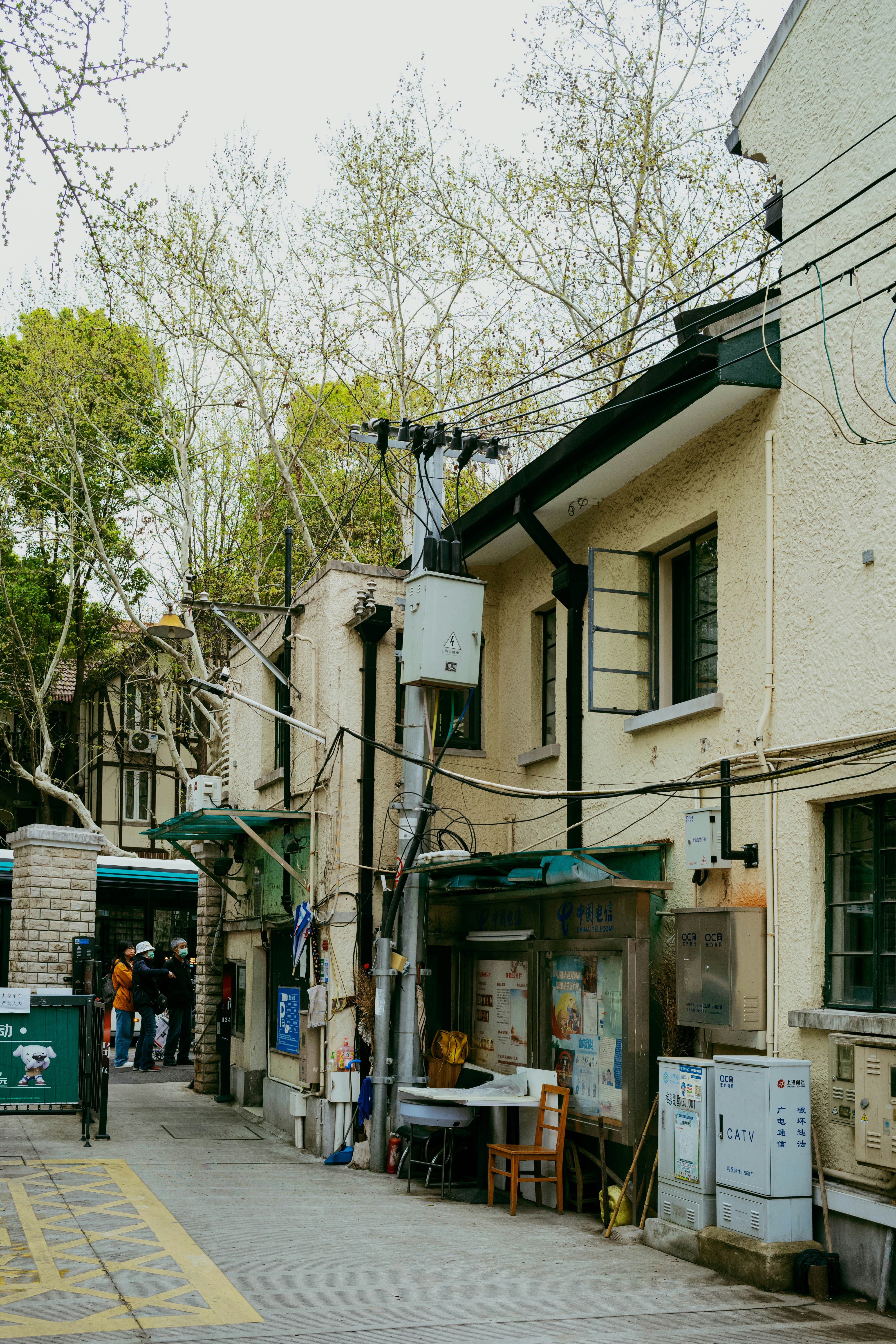 Narrow alley with rustic buildings and greenery overhead, leading to a small gathering spot.