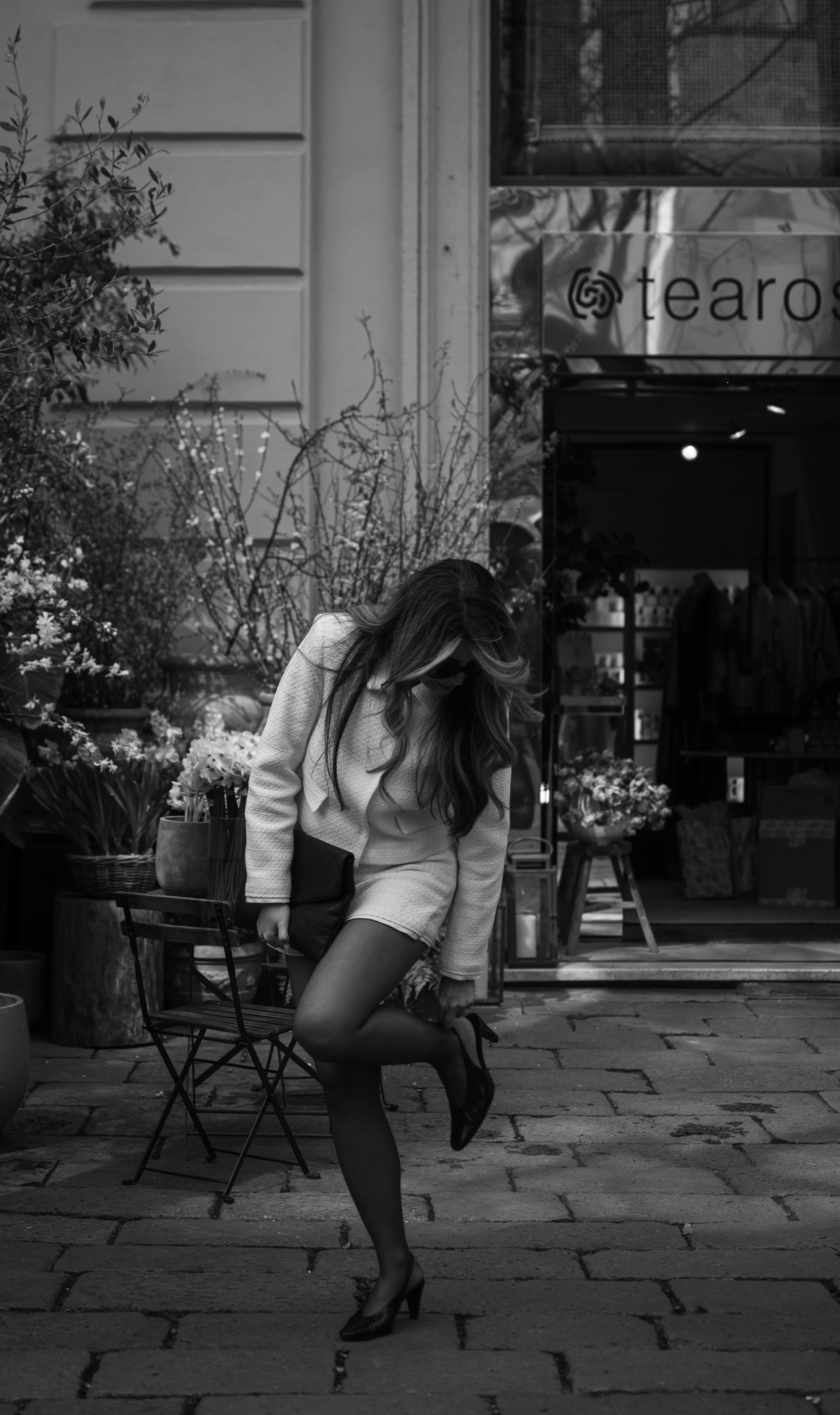 A woman adjusting her shoe outside a boutique, framed by lush greenery and stylish decor. The scene captures a moment of urban fashion in a lively setting.