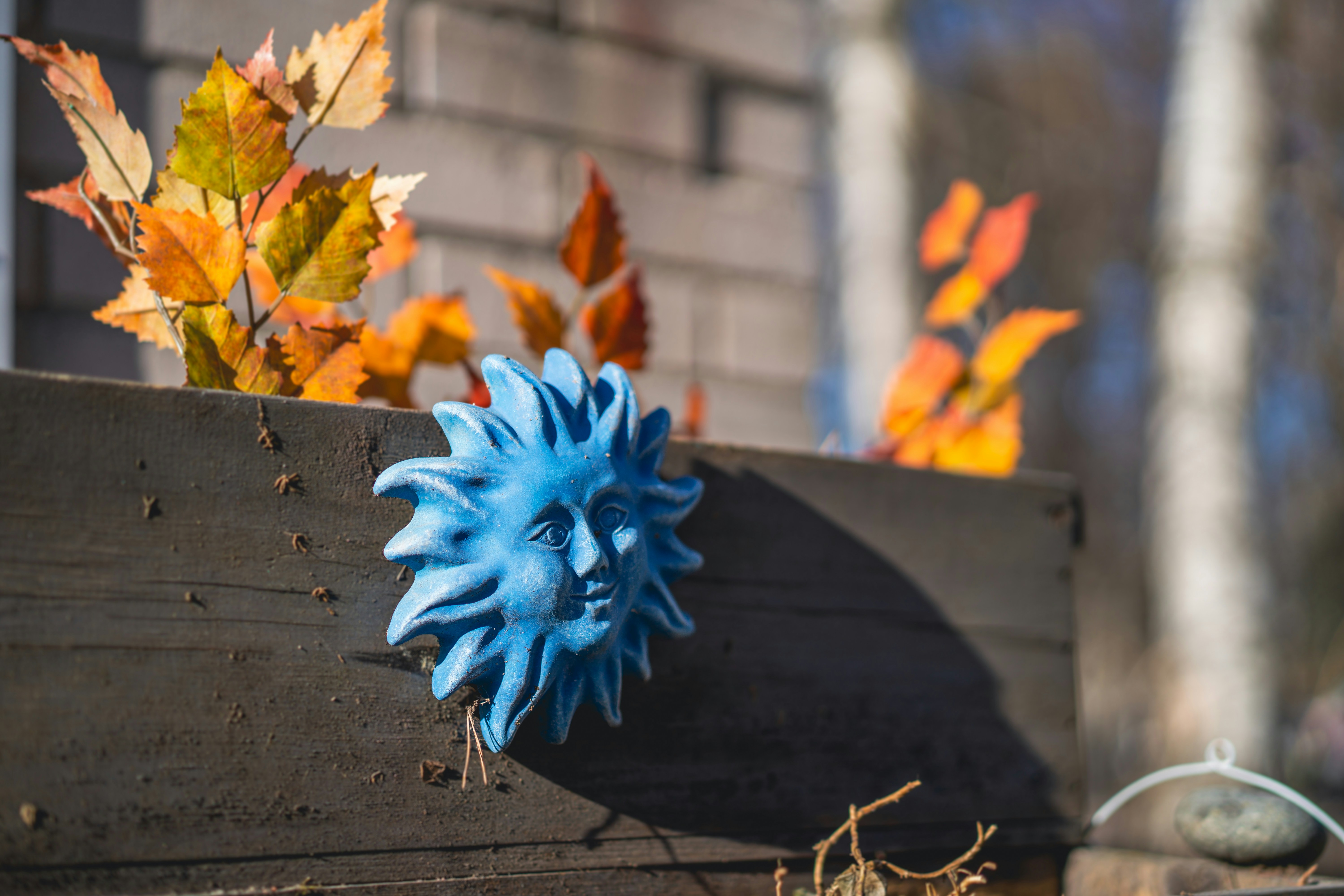 Bright blue sunface sculpture on a wooden planter with vibrant autumn leaves.