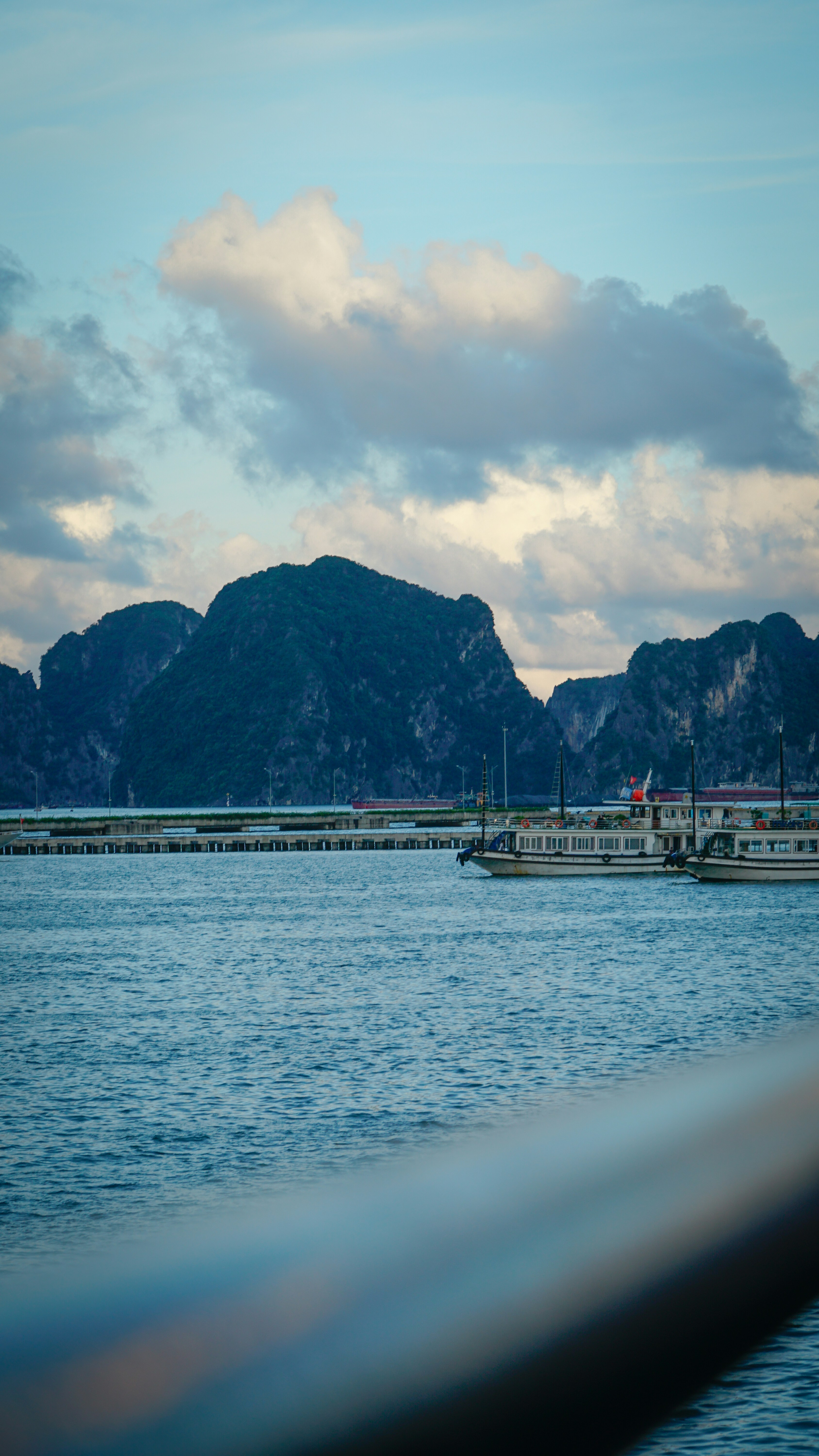 Boats docked by a tranquil sea with distant rugged mountains under a cloud-speckled sky.
