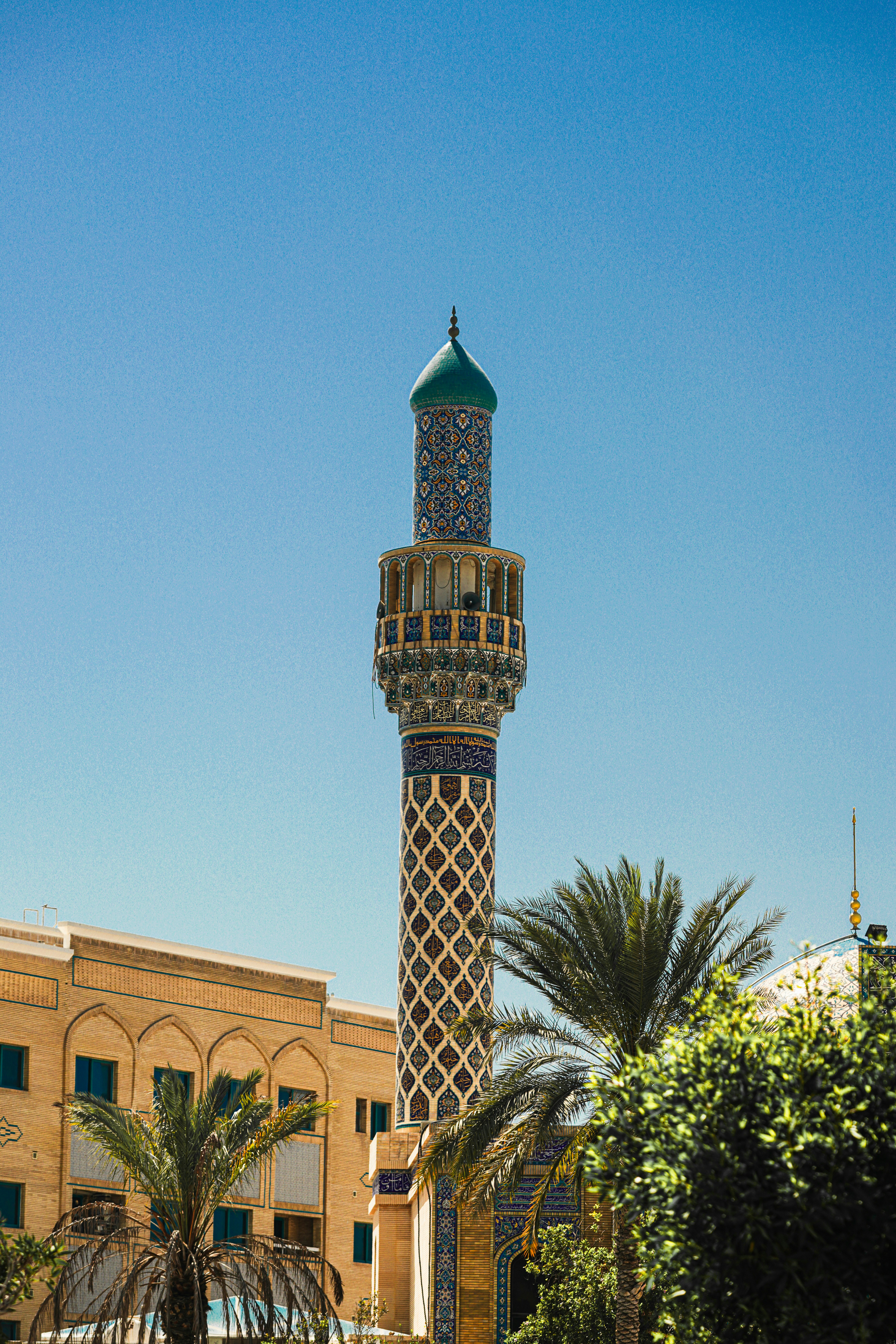 Intricate minaret adorned with vibrant tiles stands tall amidst palm trees and clear blue skies.