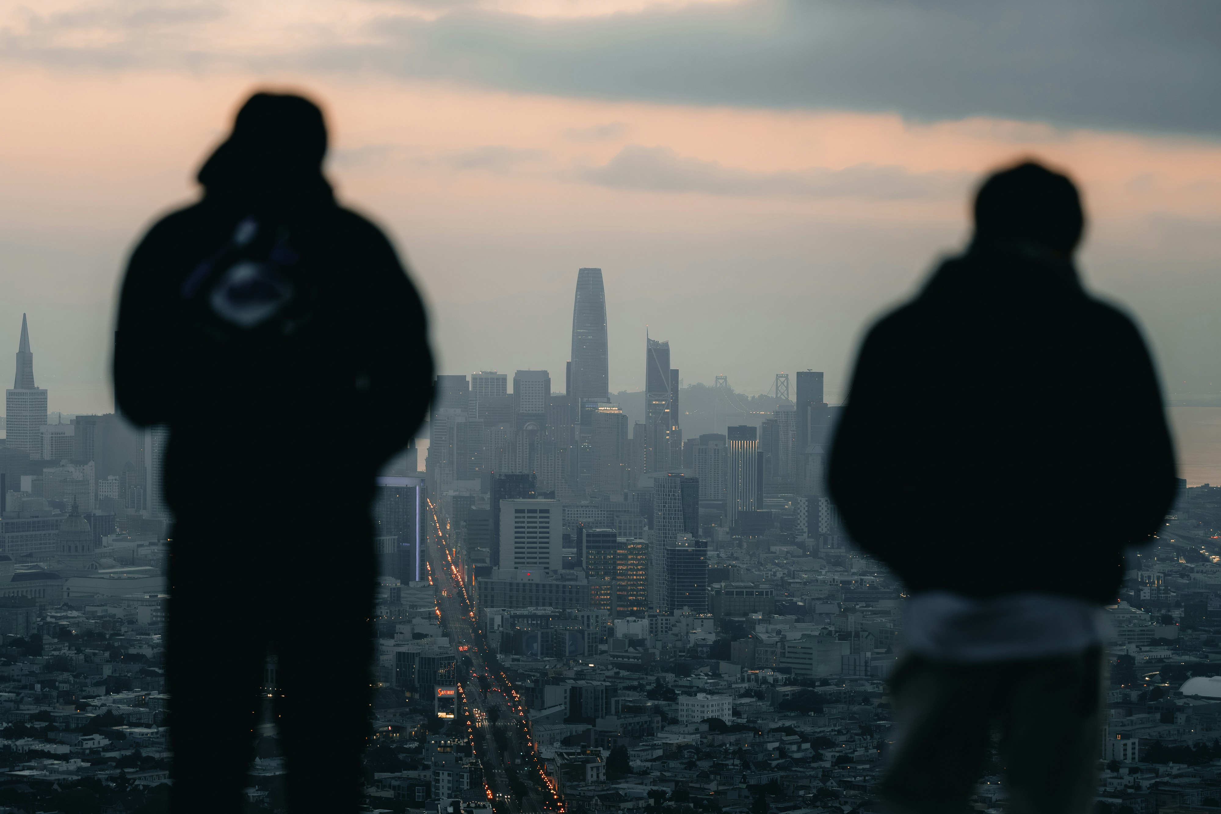 Two silhouetted figures overlook a sprawling cityscape at dusk, with towering skyscrapers in the distance.