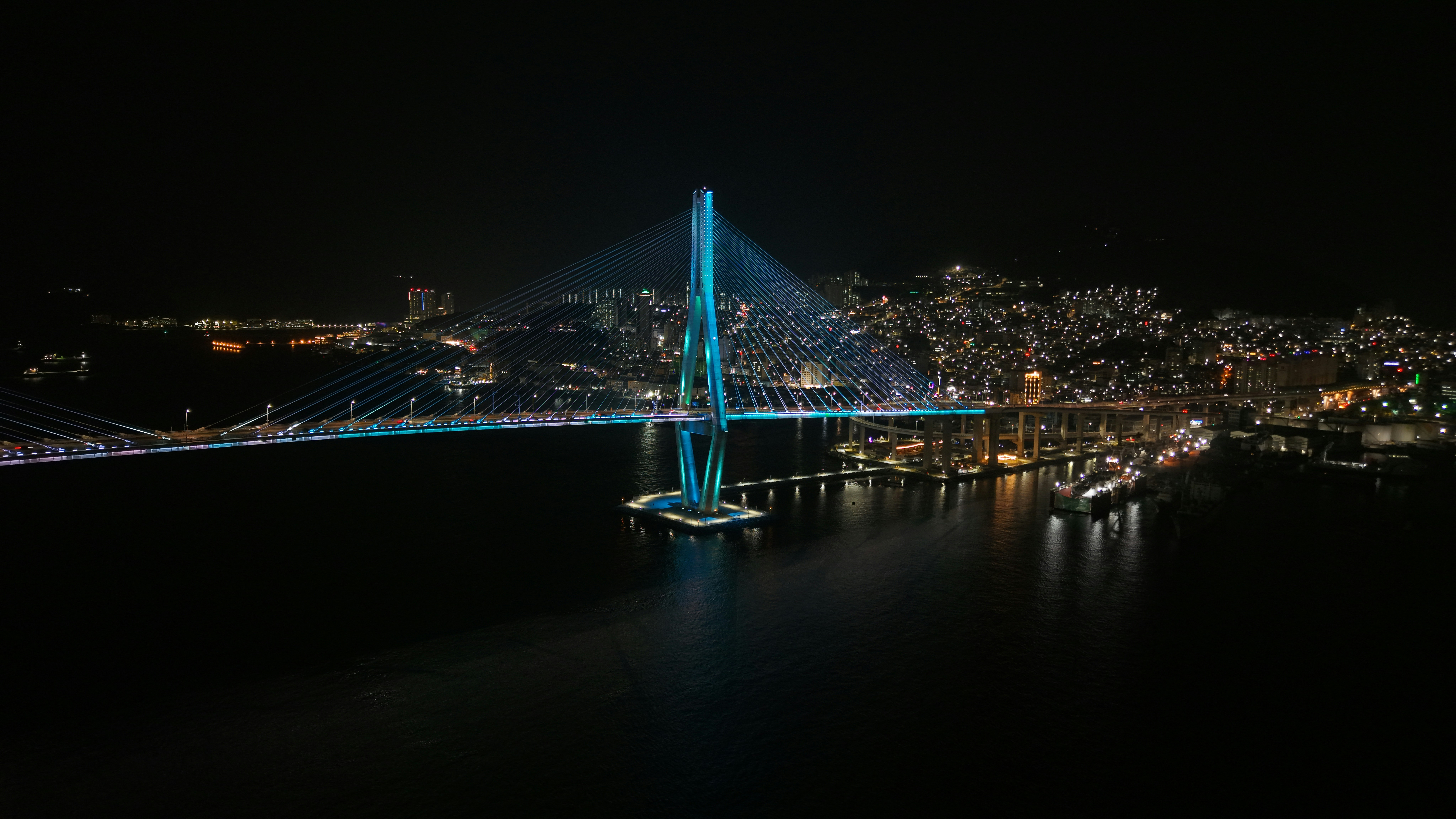 Bridge adorned with vibrant blue lights spanning a river against a backdrop of city lights at night.