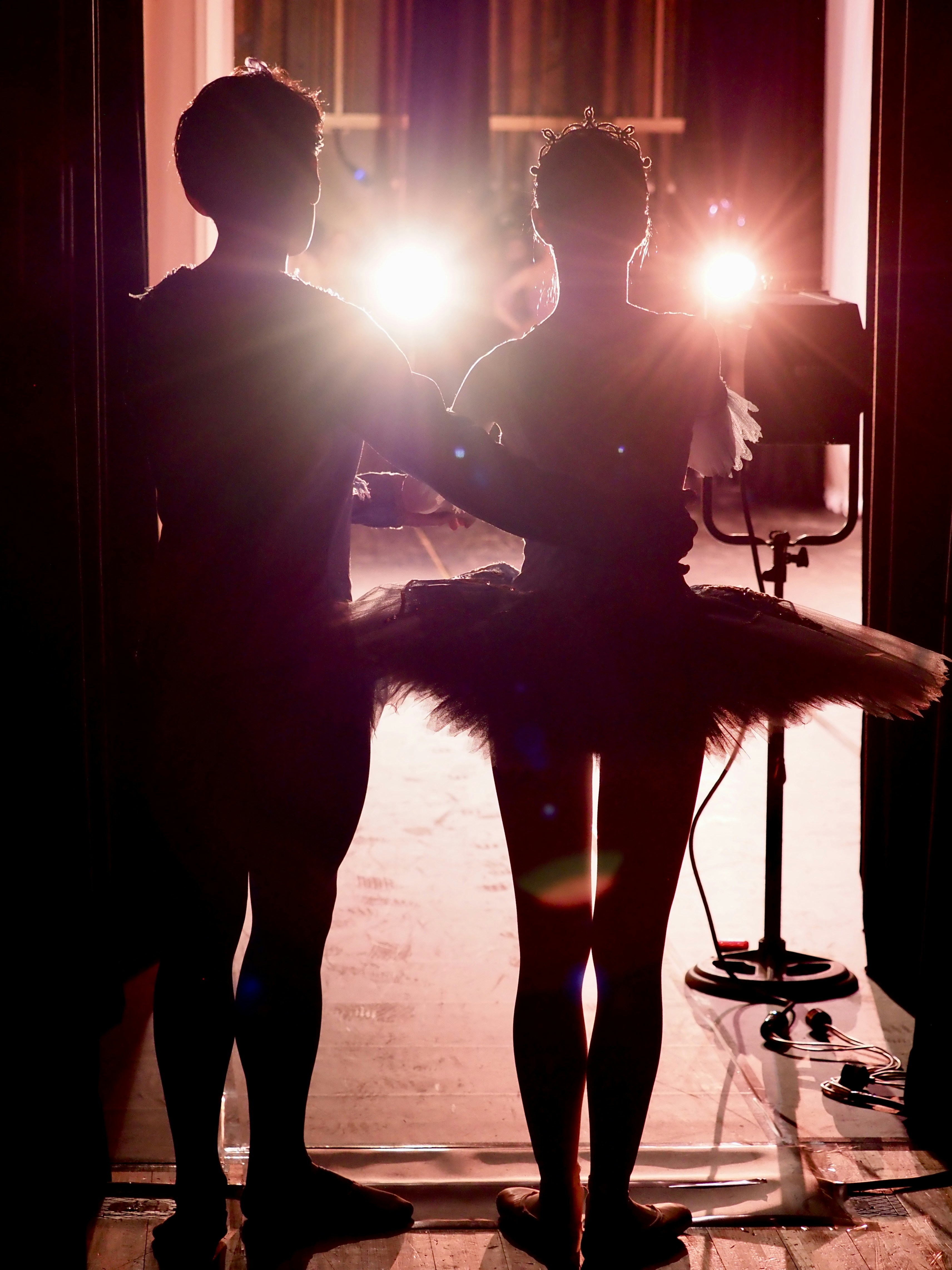 Silhouetted dancers stand poised backstage, illuminated by stage lights.