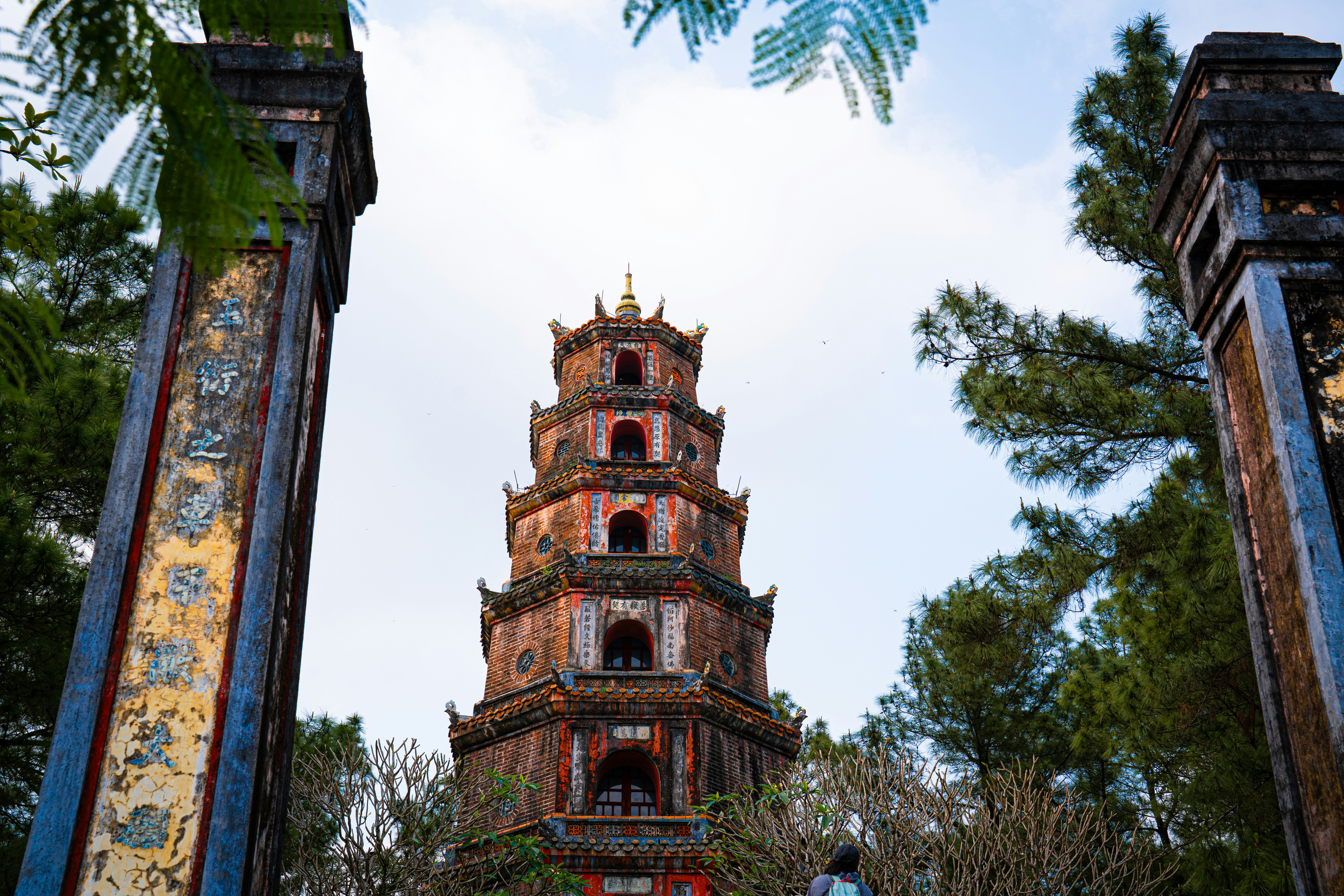 Tall pagoda rises between decorative pillars under a bright sky, surrounded by lush greenery.