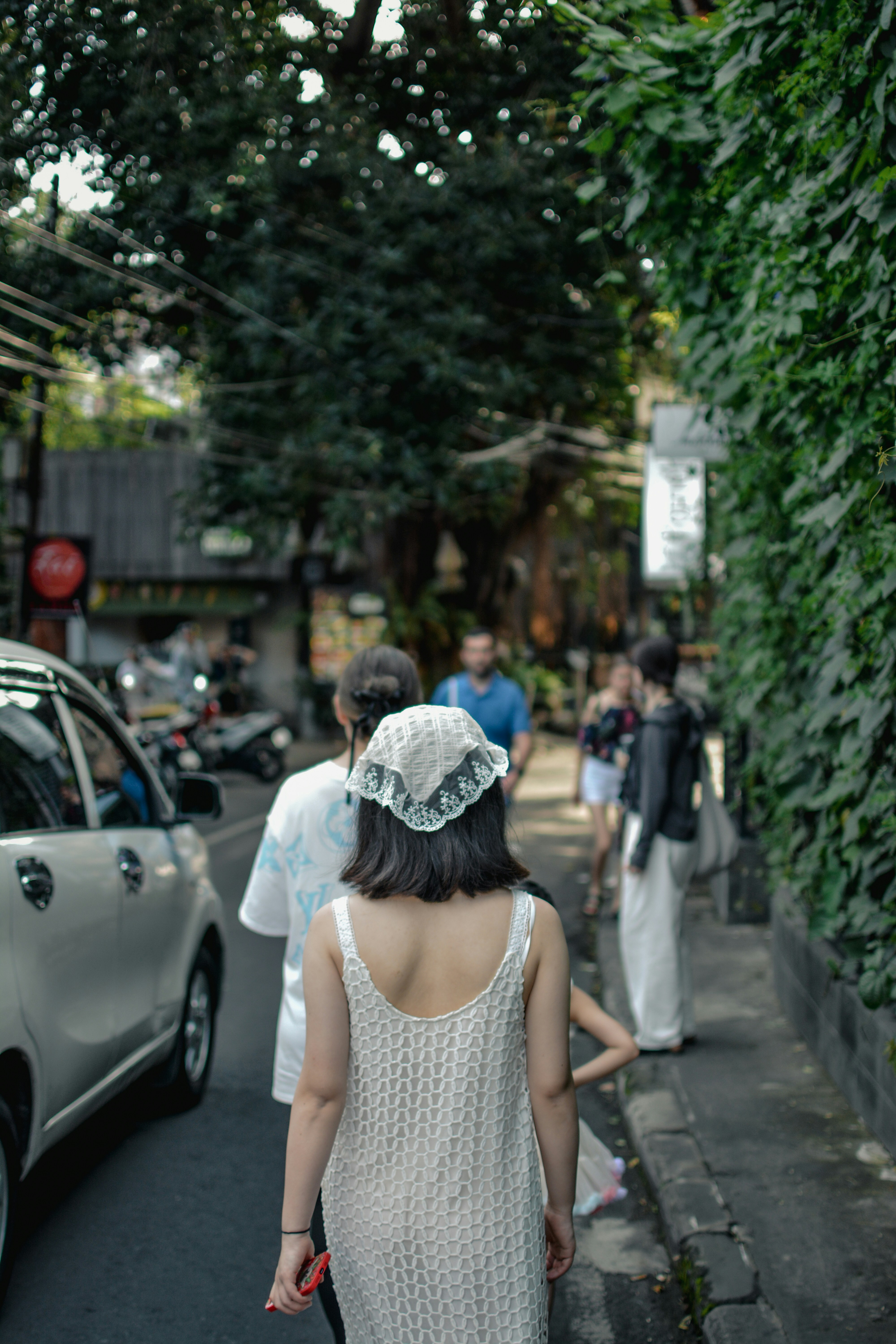 Person in a white dress and hat walking along a leafy street with cars and pedestrians.
