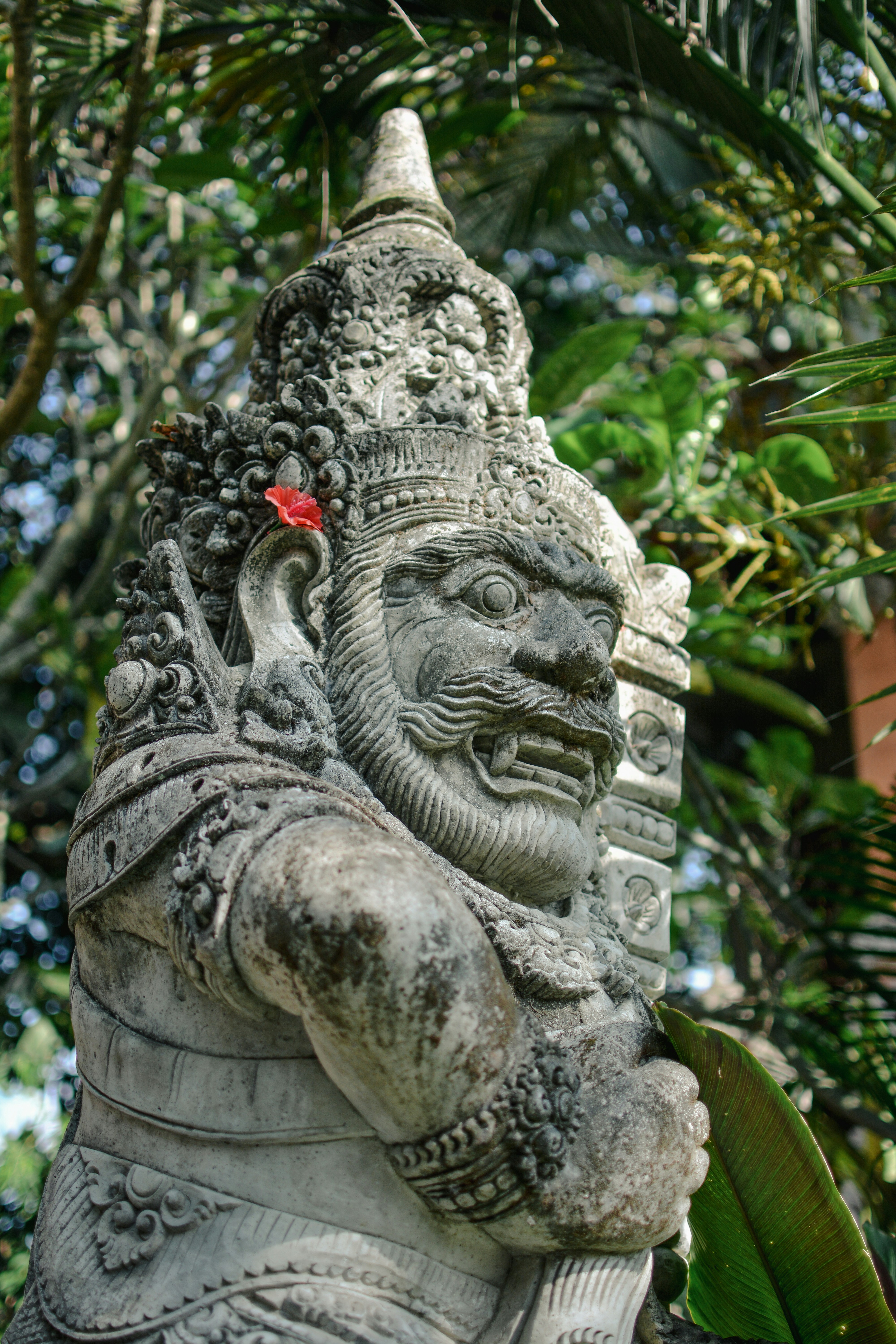 Stone statue of a mythical creature adorned with intricately carved details and a red flower, set against lush greenery.