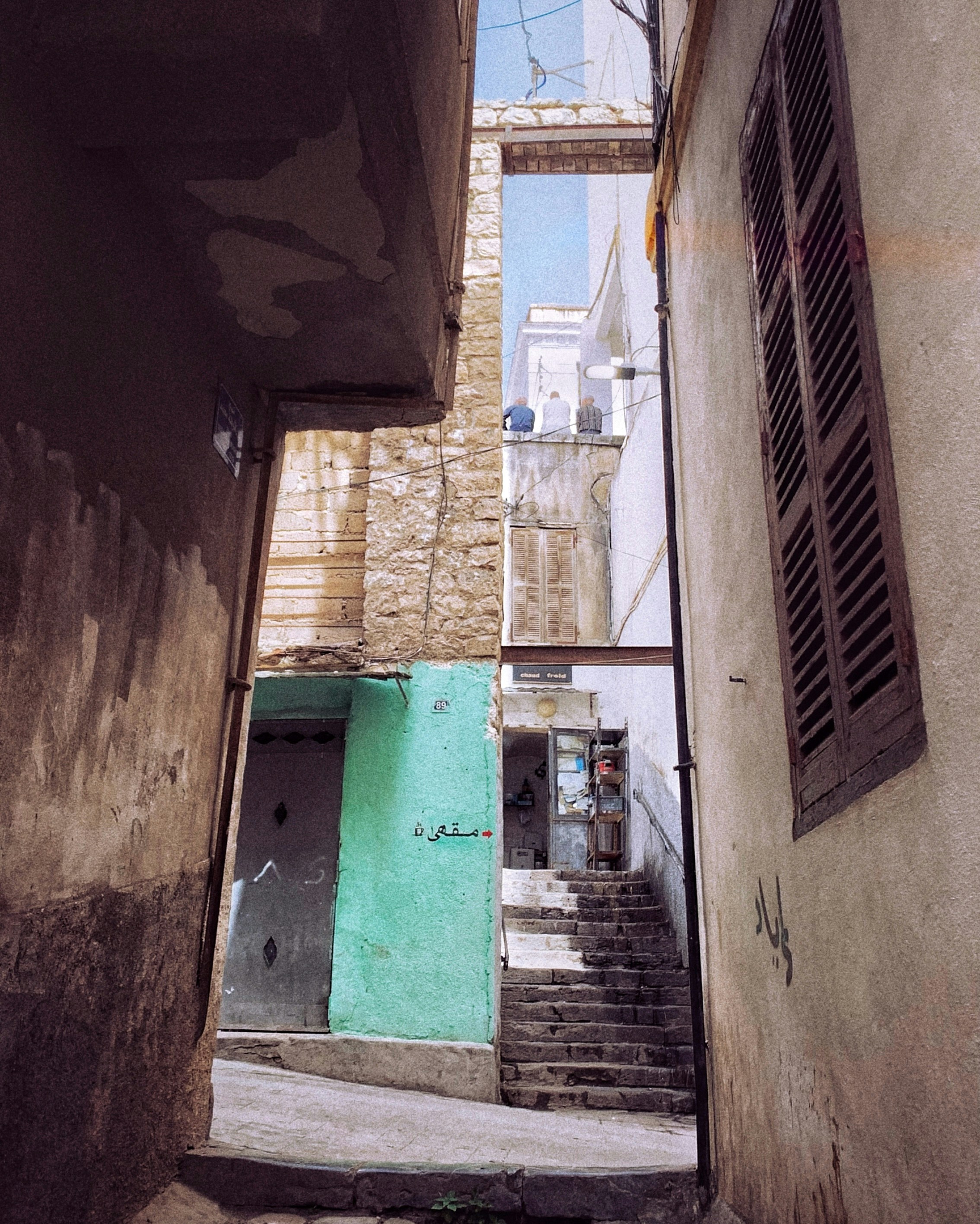 Narrow alleyway leading to a sunlit staircase with weathered walls and a green-painted facade.