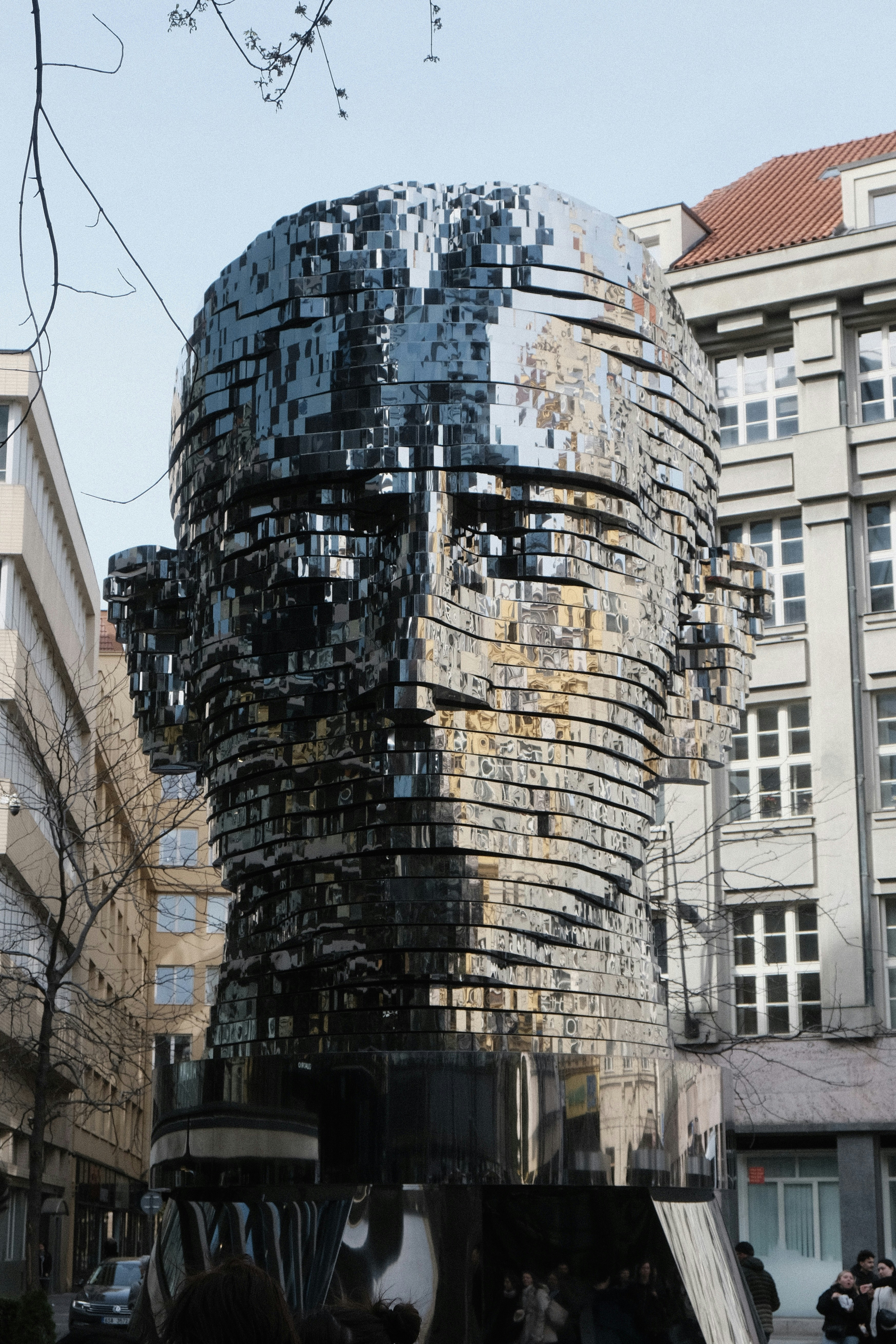 Kinetic mirrored head sculpture set against an urban backdrop of multi-story buildings under a clear sky.