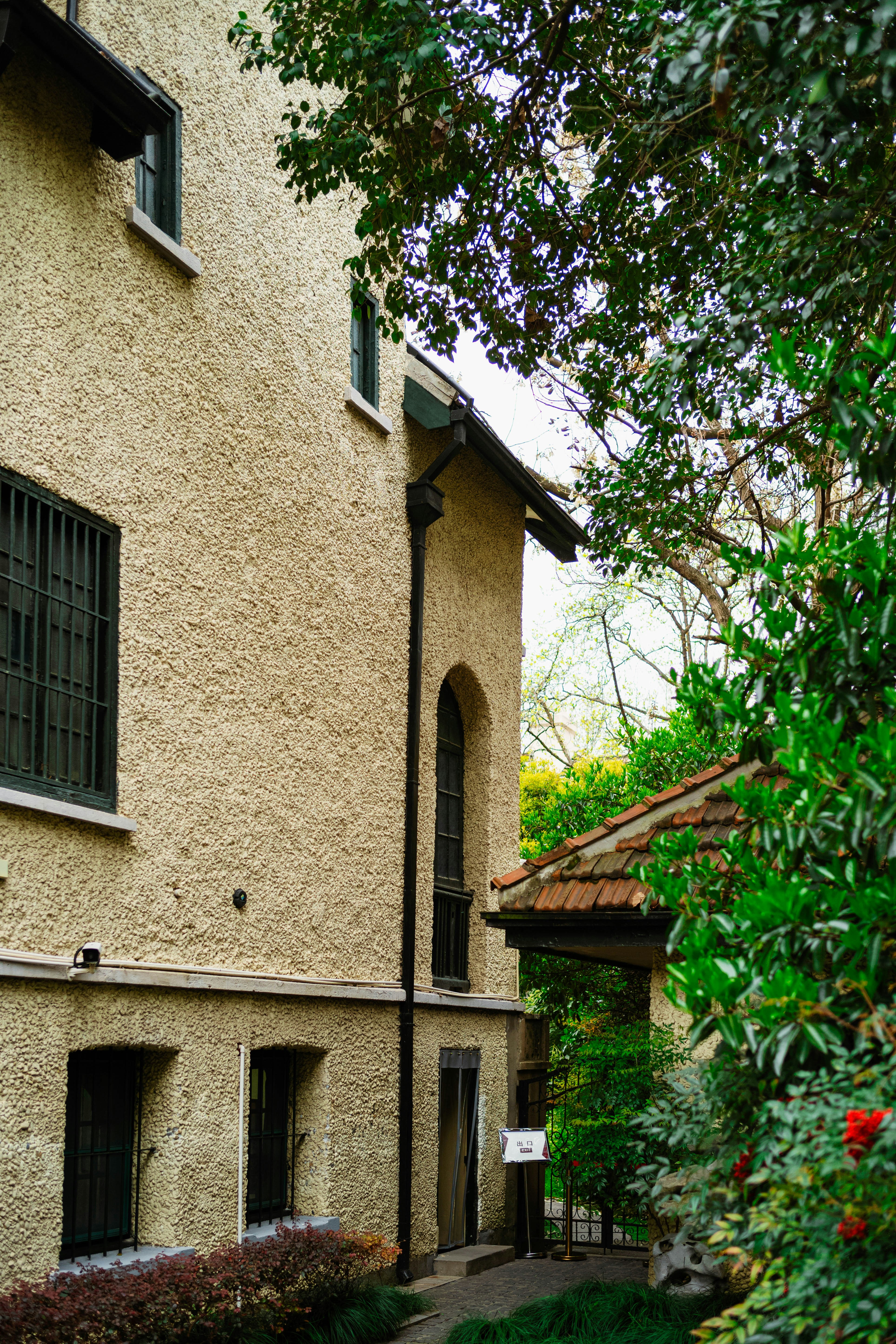 Stucco building with arched window beside a lush garden path under a cloudy sky.