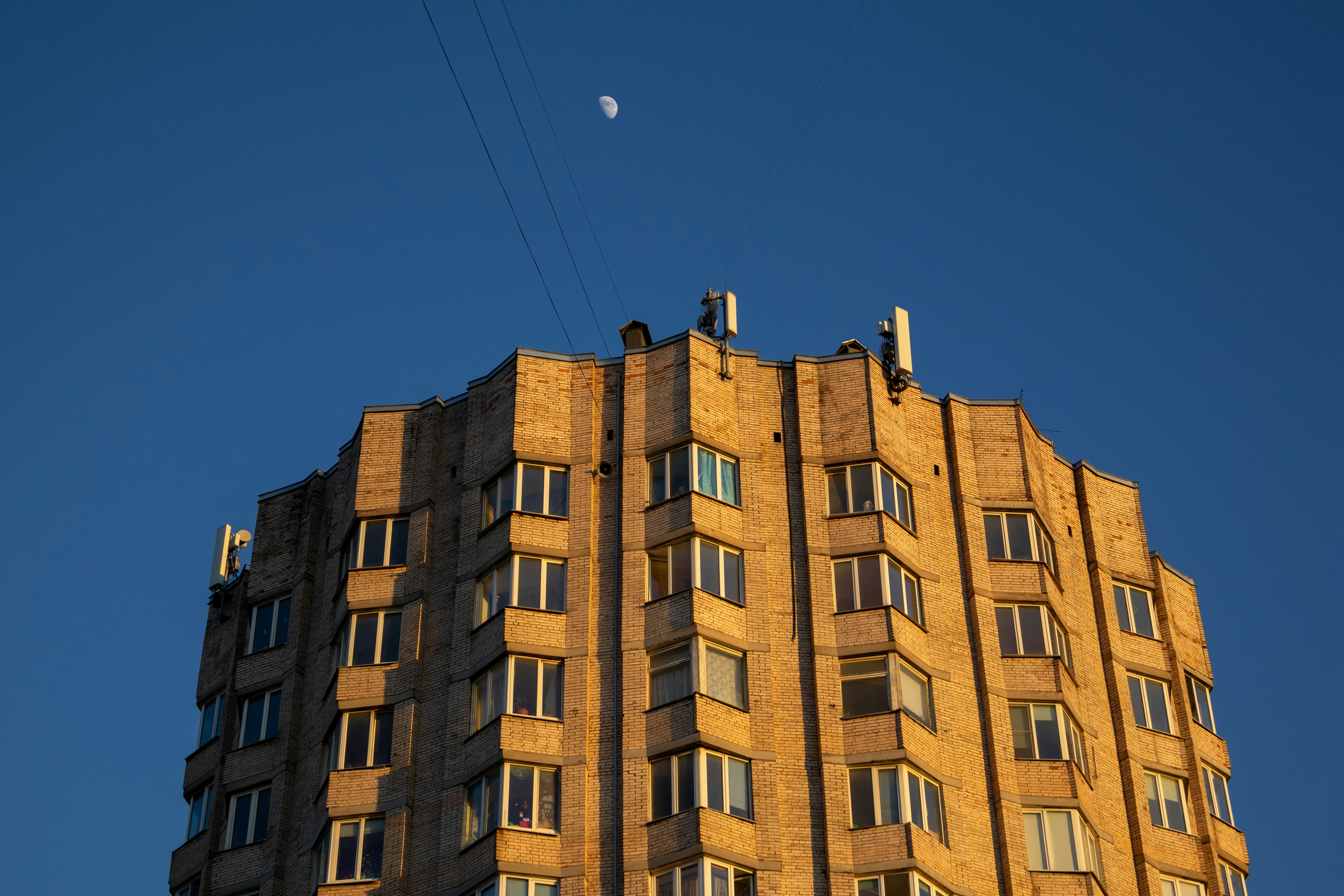 A tall, angular building bathed in warm light contrasts sharply with a clear blue sky and a visible crescent moon above.