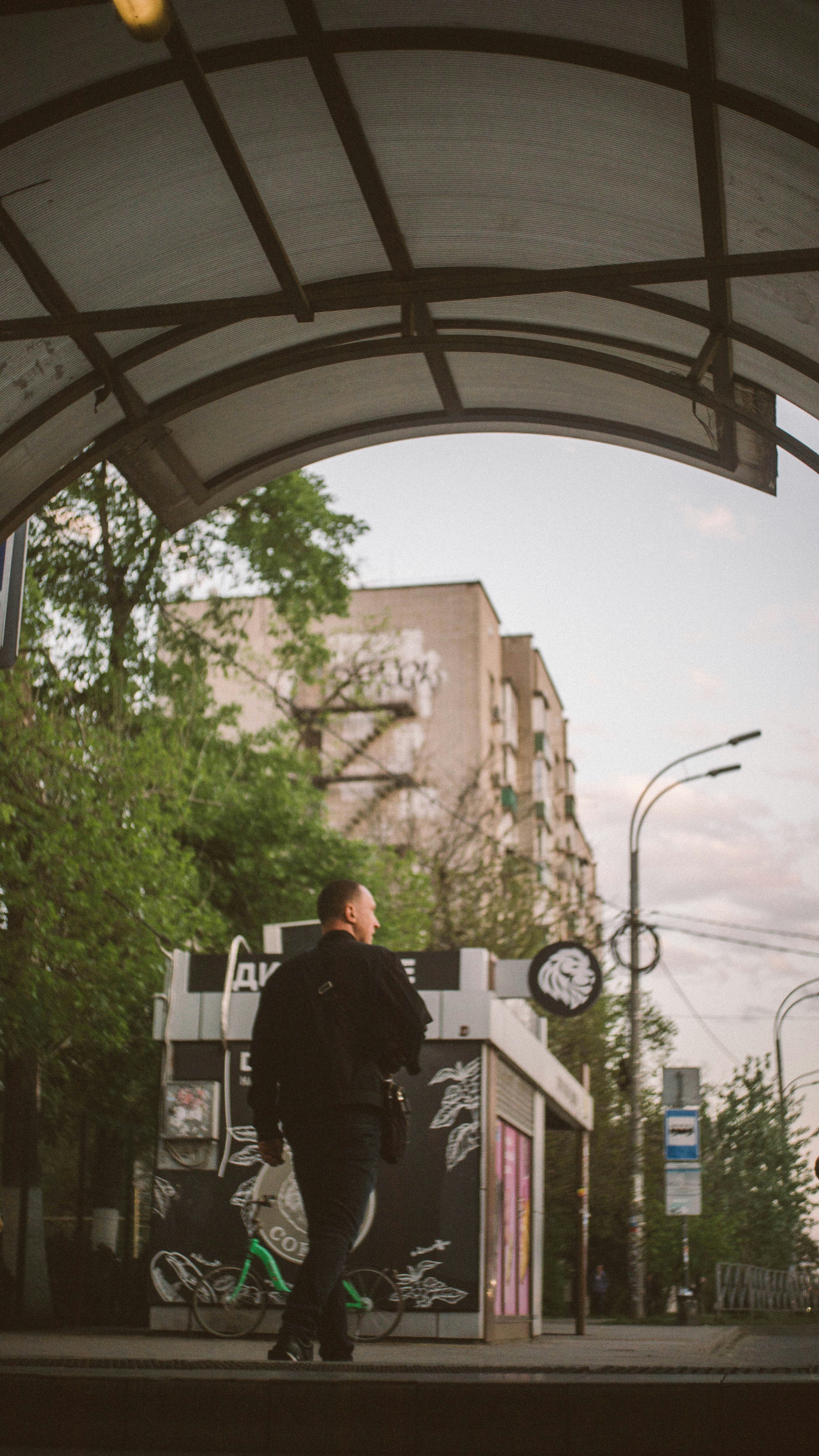 Person walking under a curved canopy with urban buildings and trees in the background.