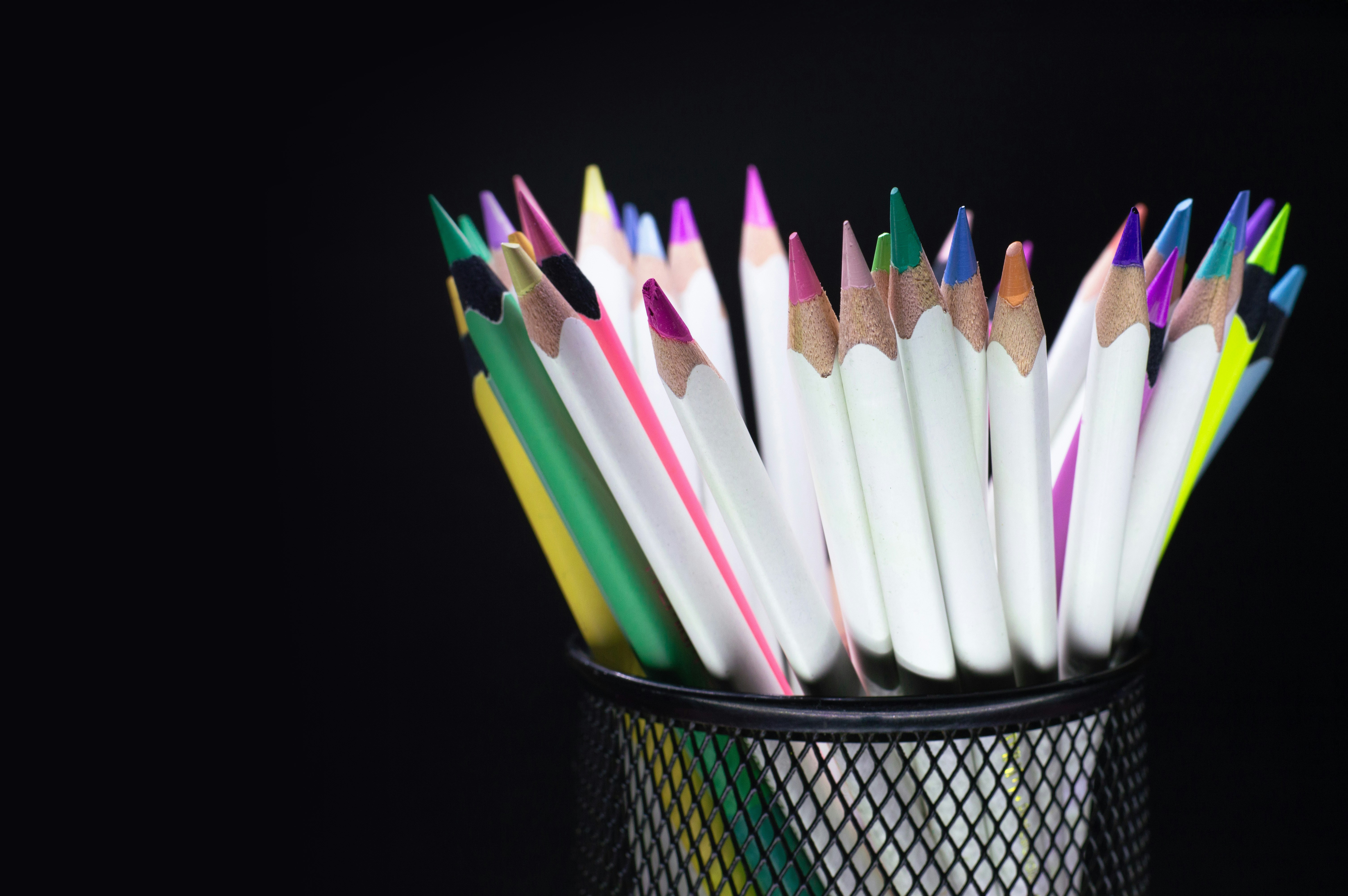 Assorted colored pencils in a black mesh holder against a dark background.