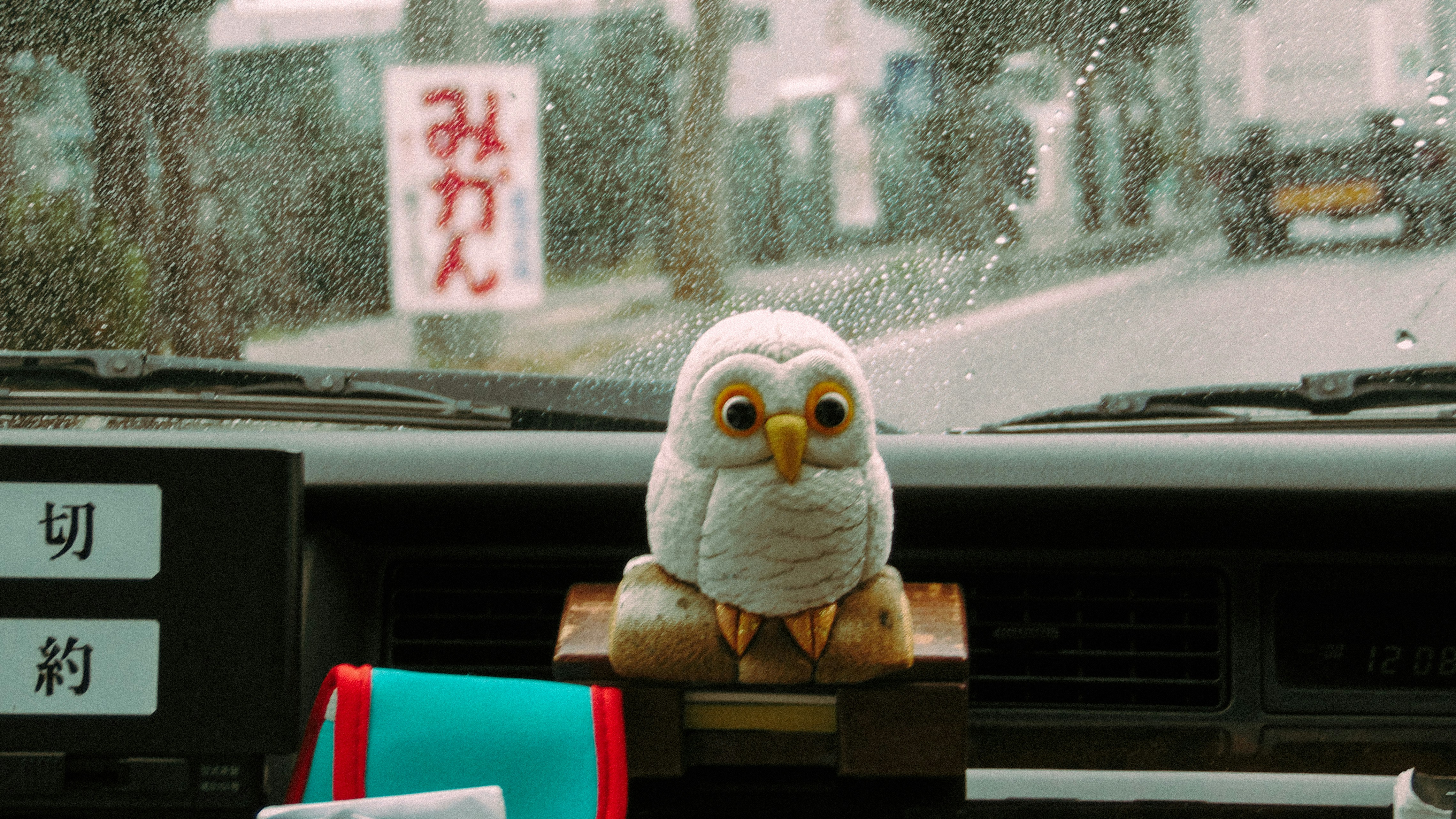Plush owl figurine perched on a taxi dashboard, framed by a rain-speckled windshield and city street view.