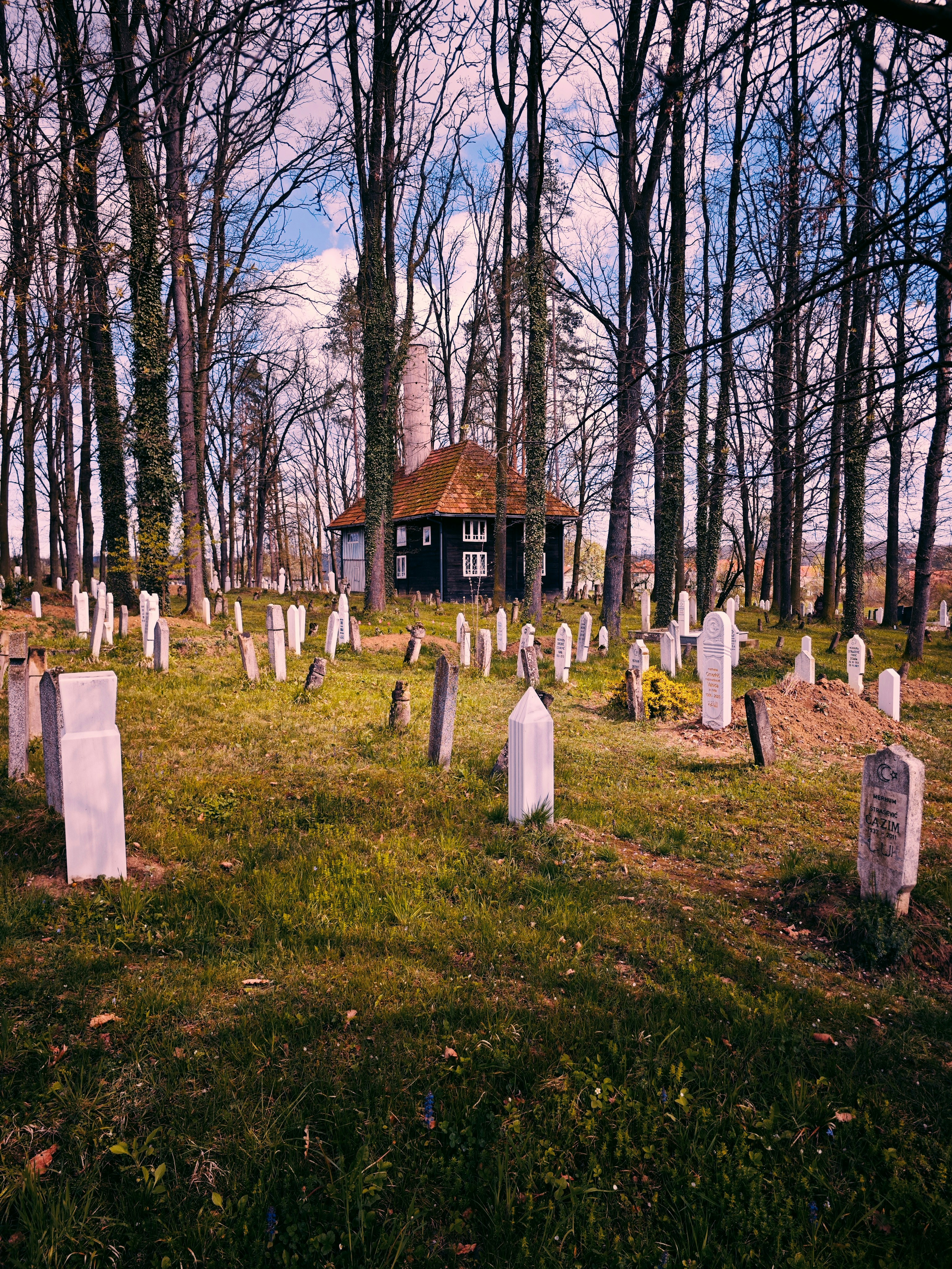 A serene cemetery surrounded by tall trees, featuring white gravestones and a quaint black house in the background. The scene evokes a sense of tranquility and reflection.
