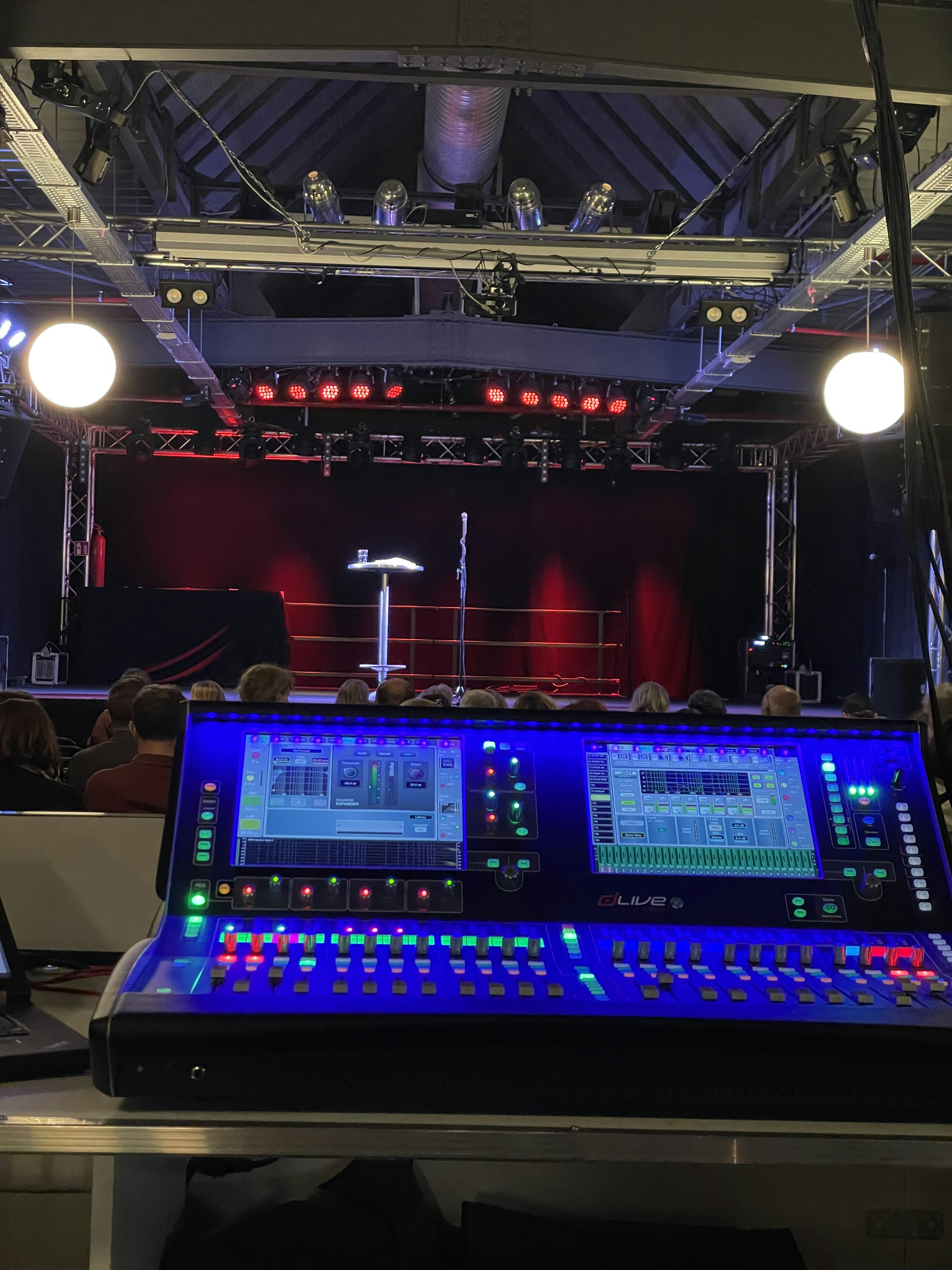 Mixing console glowing in vibrant colors in front of an empty stage with red and blue lighting in a concert venue.