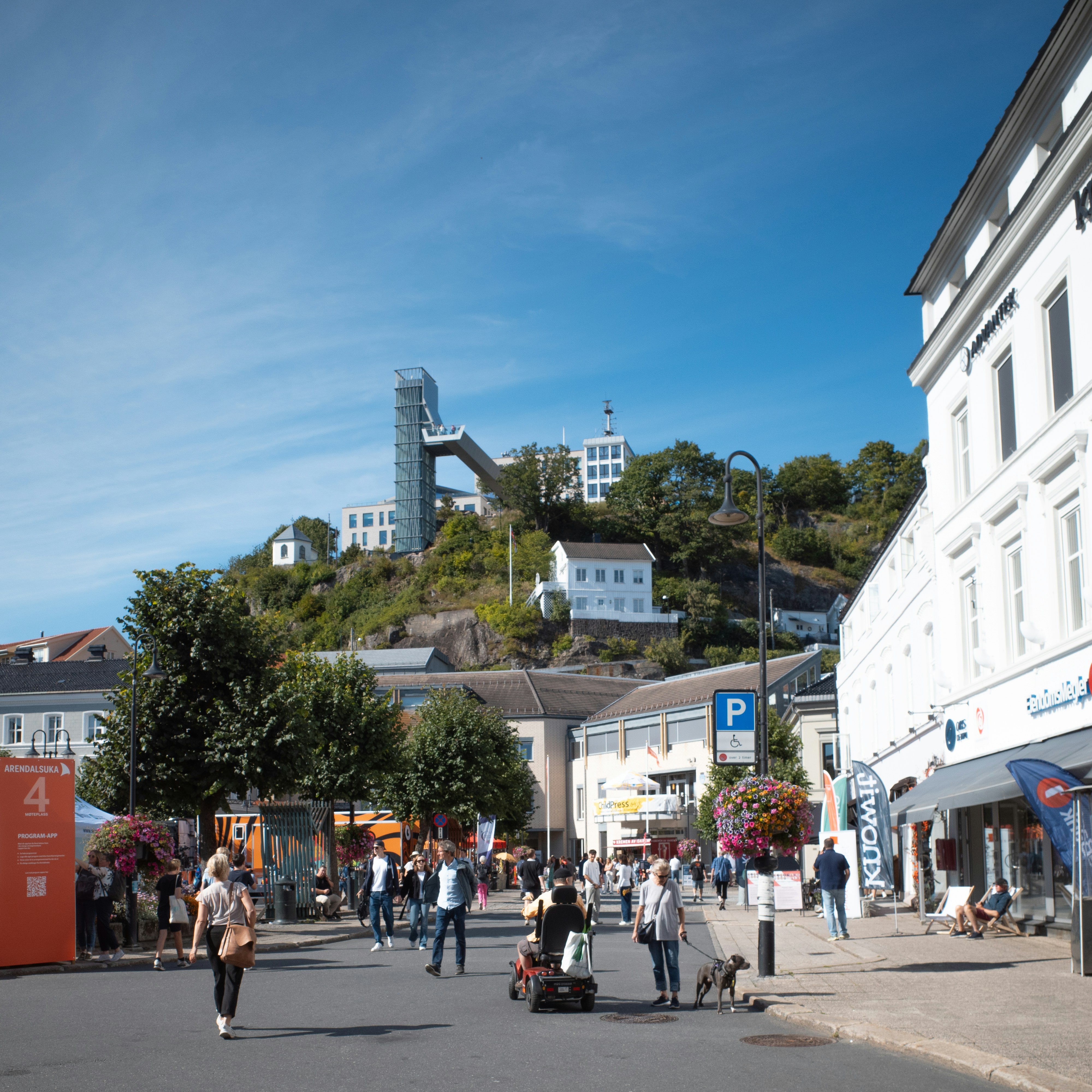 Busy street scene with people strolling, set against a backdrop of a hilltop building and a blue sky.