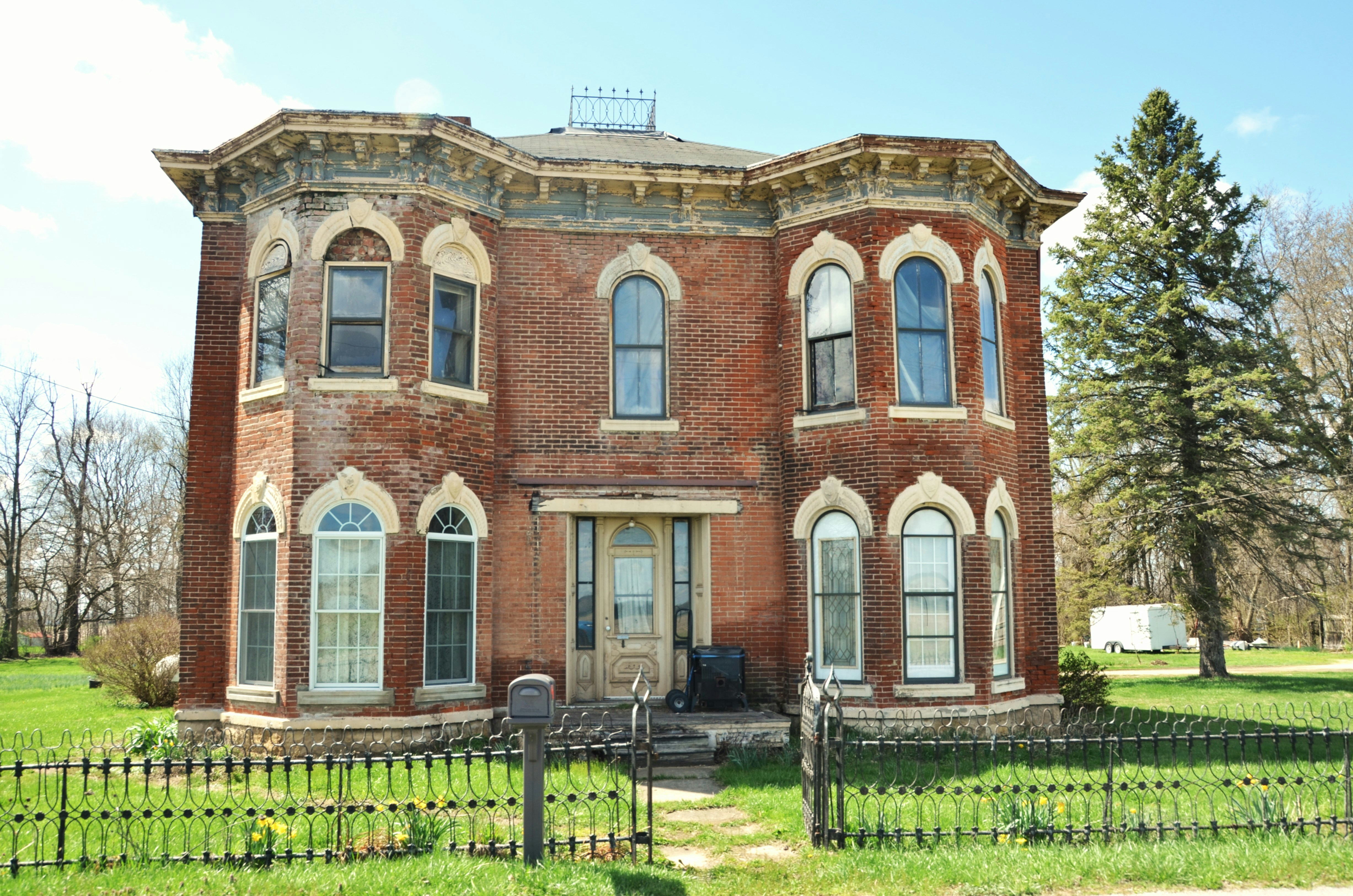 Historic brick farmhouse with unique architectural details and an ornate iron fence under a clear sky.