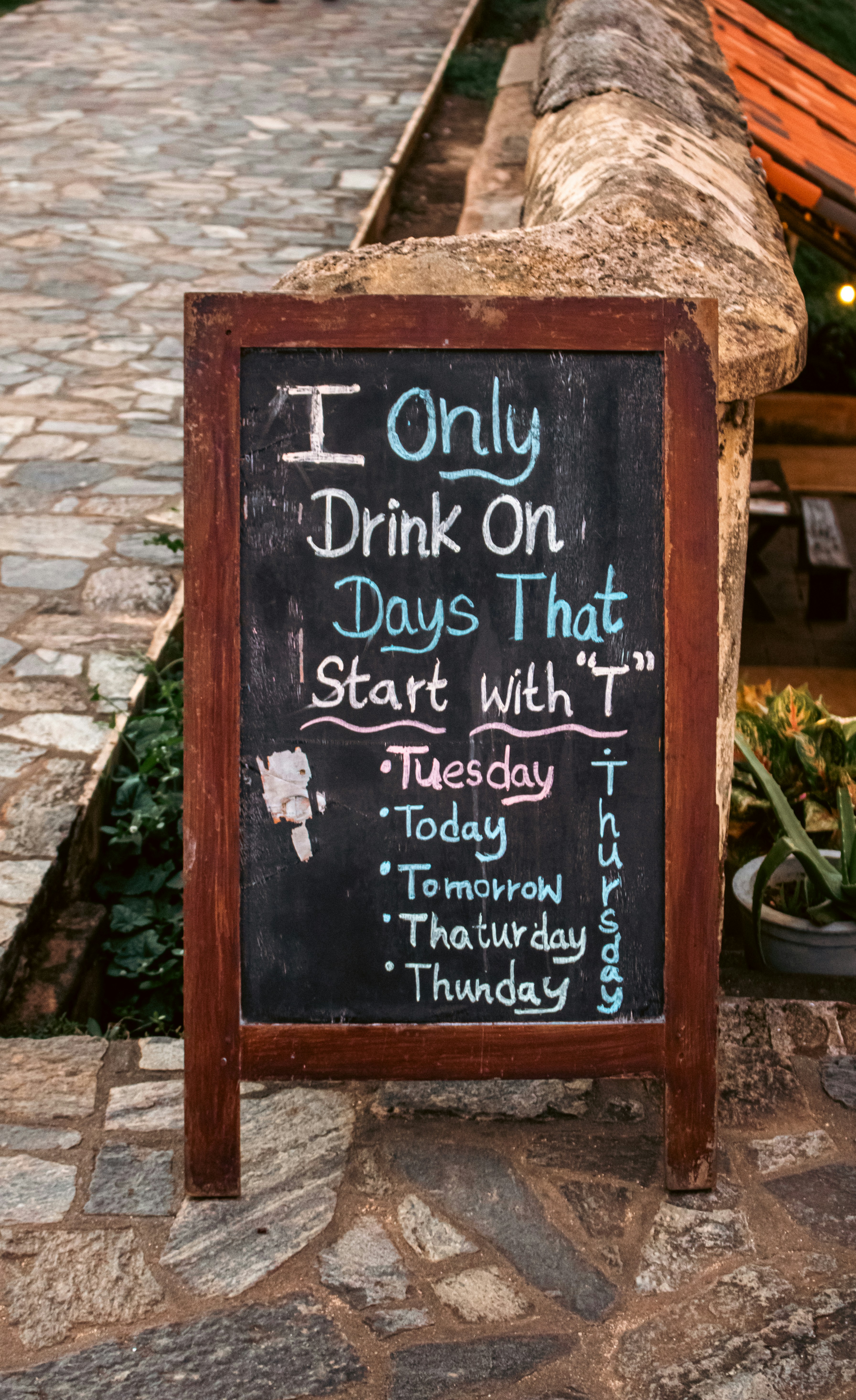 A wooden A-frame chalkboard sign stands on a stone-paved outdoor walkway, at the entrance to a bar and restaurant located within the Dutch Fort in Galle, Sri Lanka. Written in colorful chalk, it displays a humorous message: "I Only Drink On Days That Start with 'T'". Below, it lists "Tuesday", "Today", "Tomorrow", plus the invented "Thaturday" and "Thunday", with "Thursday" written vertically.