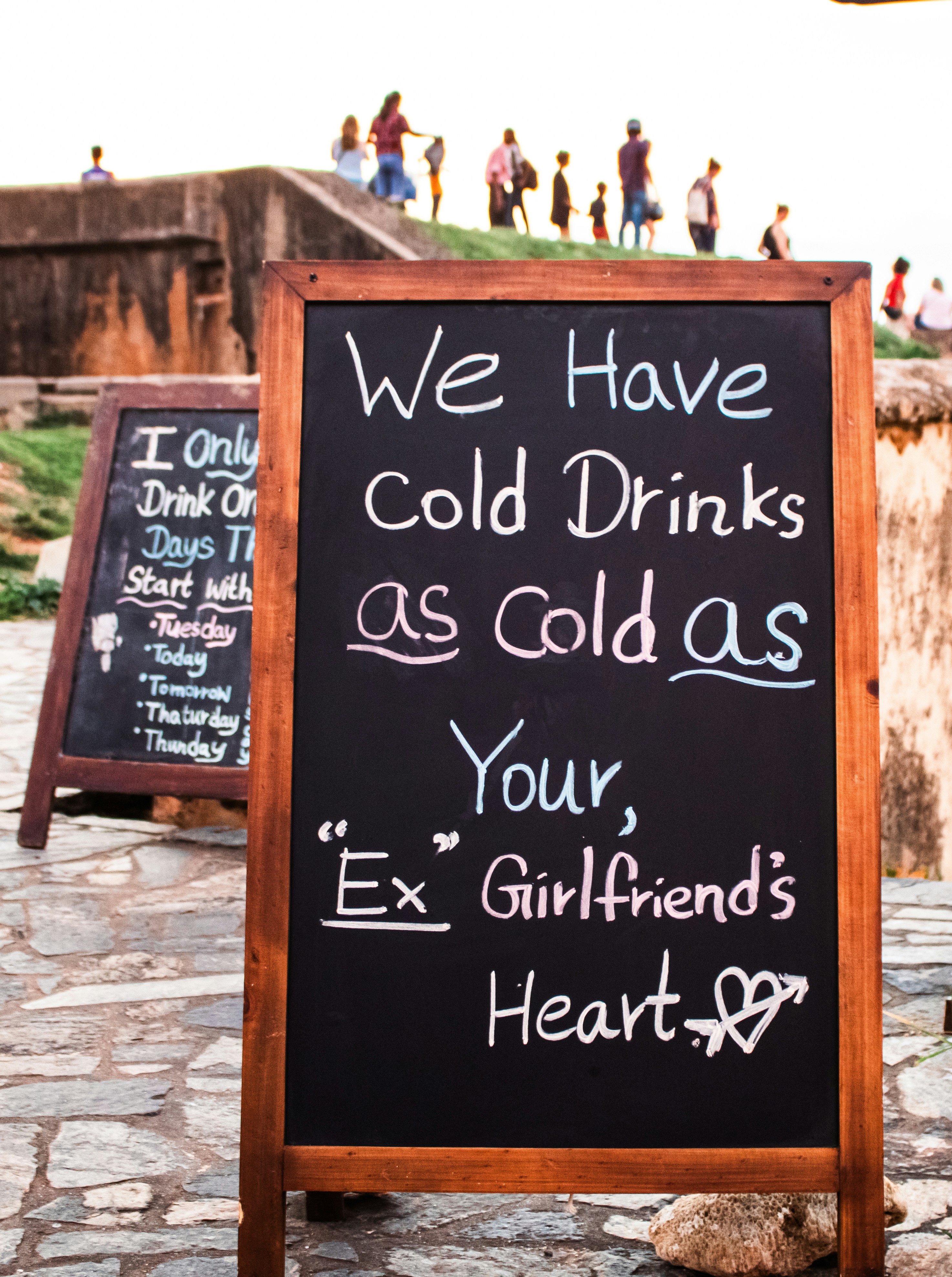 An A-frame chalkboard sign stands outside, for a pub placed on irregular stone paving. Using colorful chalk, it presents a humorous, slightly edgy advertisement: "We Have cold Drinks as Cold as Your 'Ex' Girlfriend's Heart ♡".